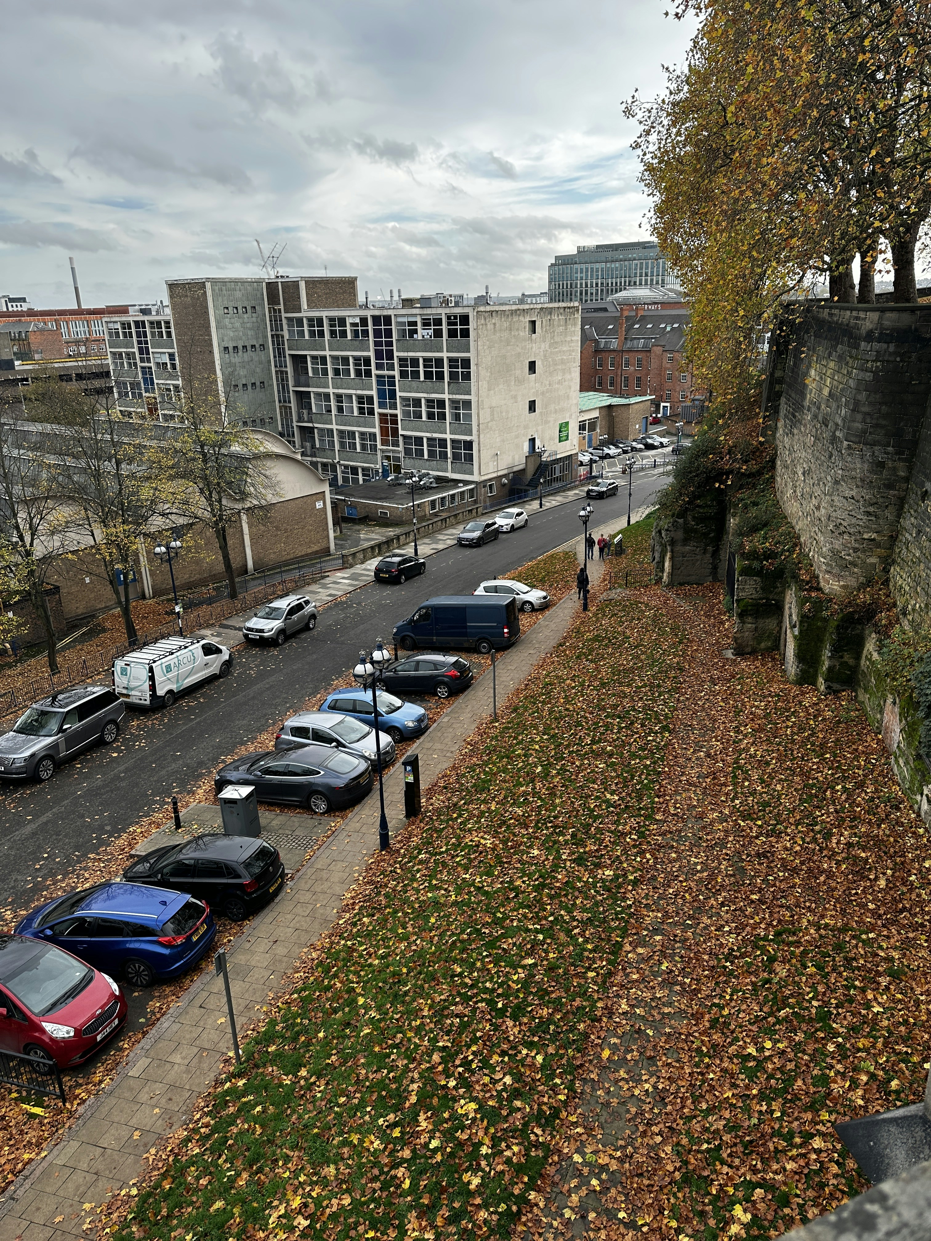 Autumn street scene with parked cars along a cobbled road beside a tree-lined slope and a high stone wall, viewed from an elevated perspective.