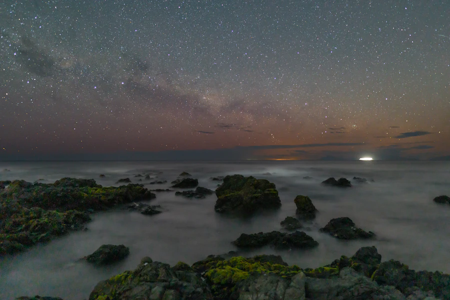 A star-filled night sky arches over a calm ocean with dark volcanic rocks in the foreground. Soft clouds and a faint glow near the horizon add color to the long-exposure scene.