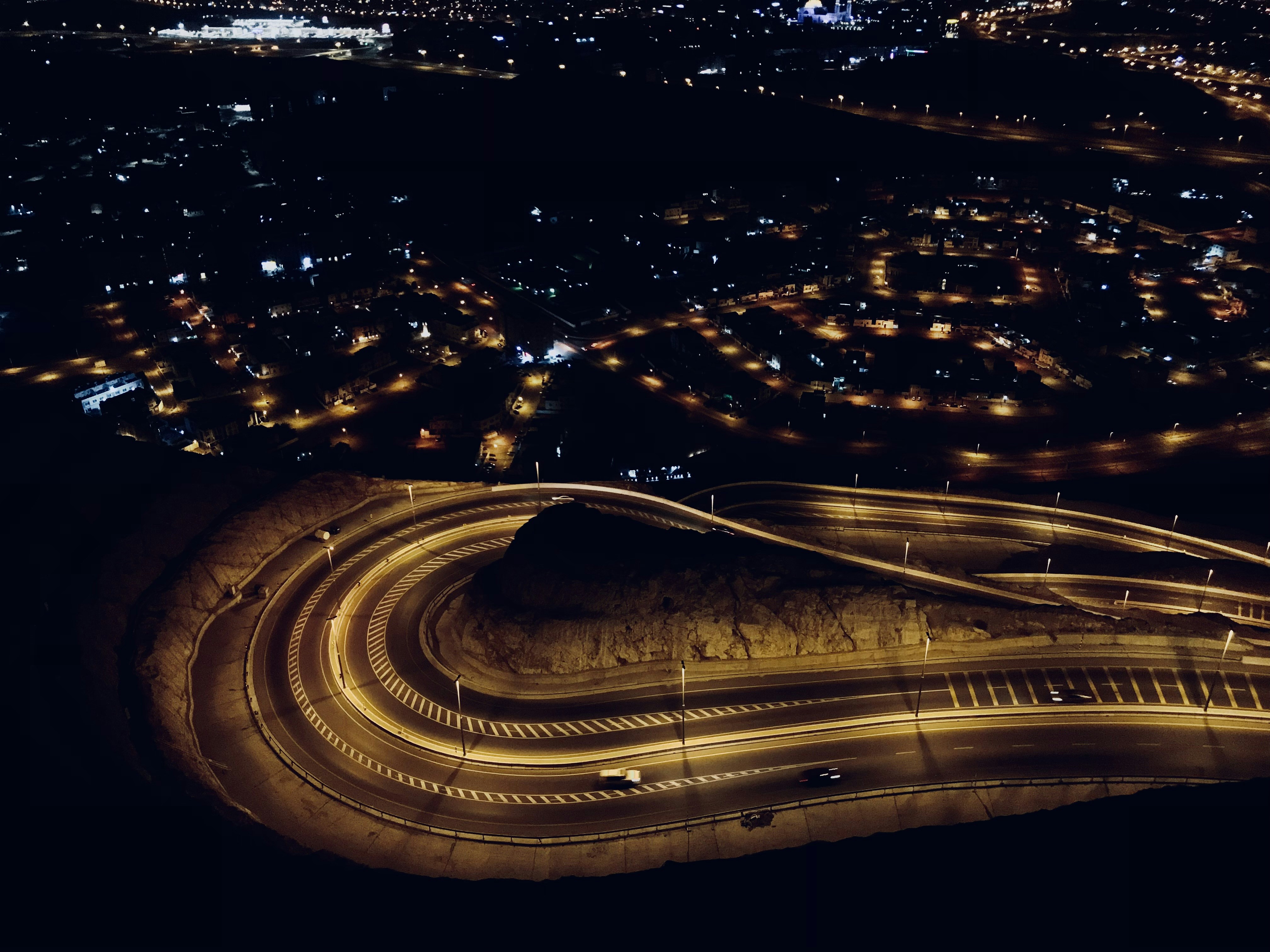Aerial view of a winding road illuminated by streetlights, showcasing the flow of traffic against a backdrop of a cityscape at night.
