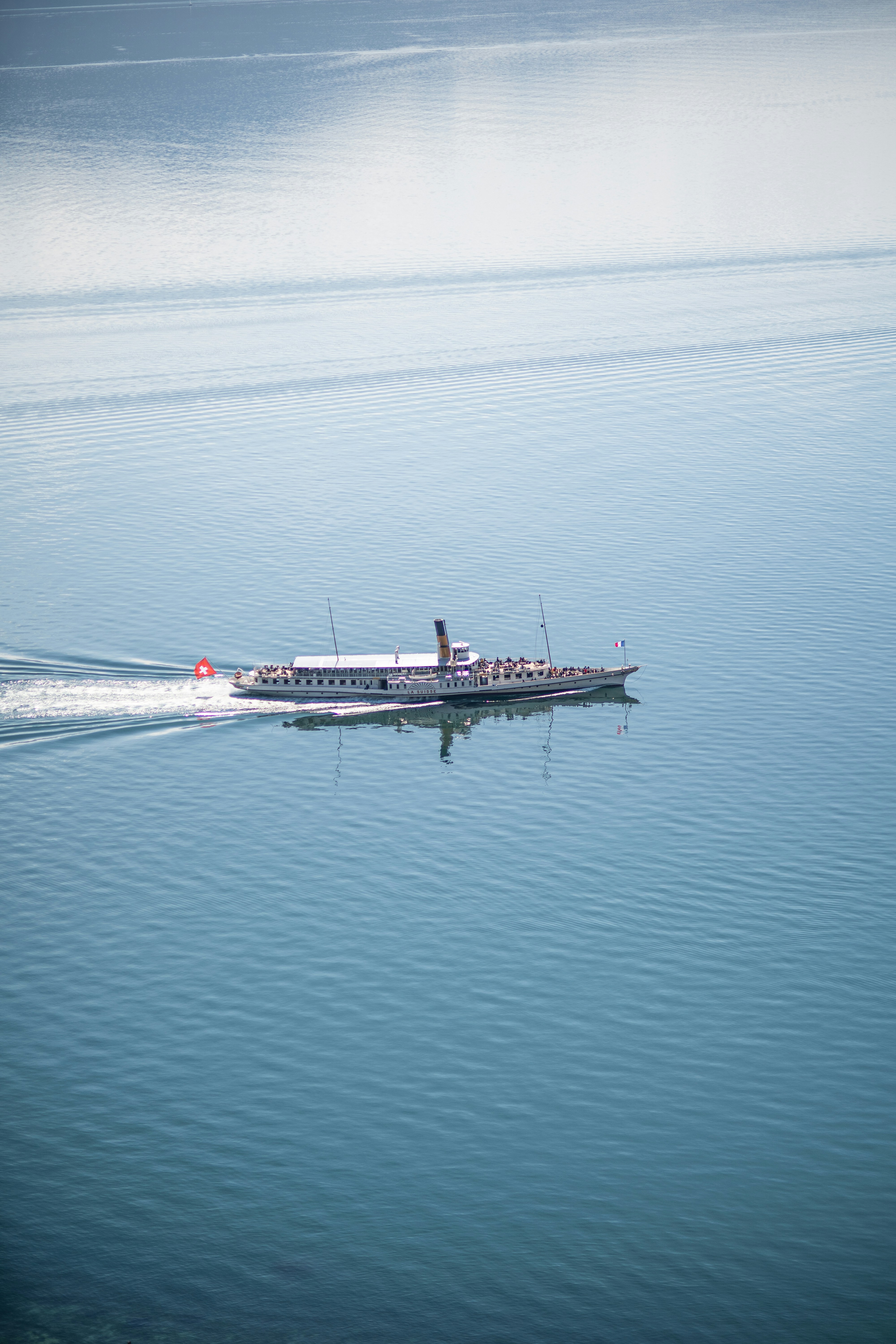 A lone steamboat glides smoothly over an expansive, calm blue lake under a clear sky.
