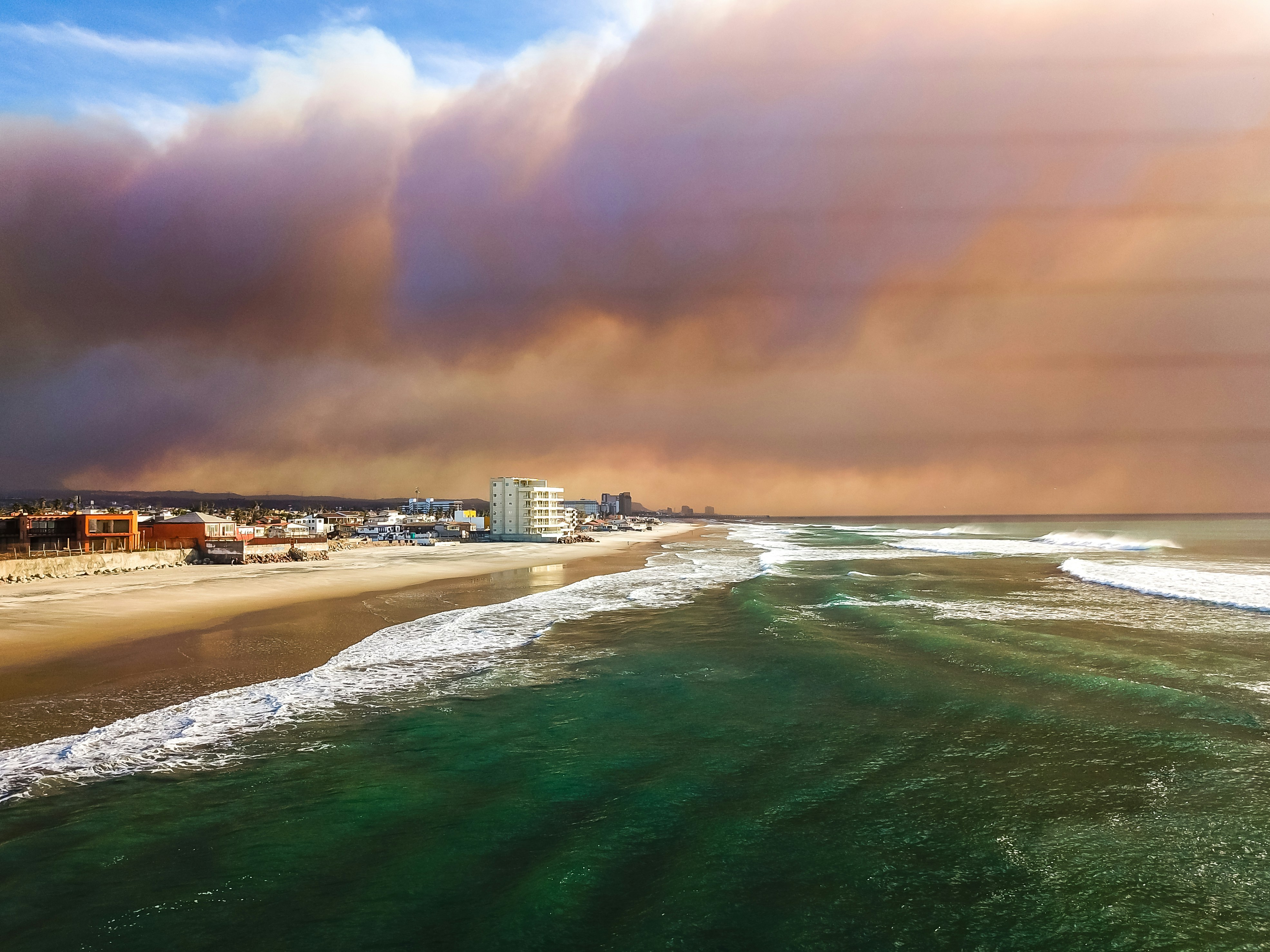 Dense smoke from wildfires drifts over Rosarito's coastline, contrasting with the vibrant ocean and clear sky.