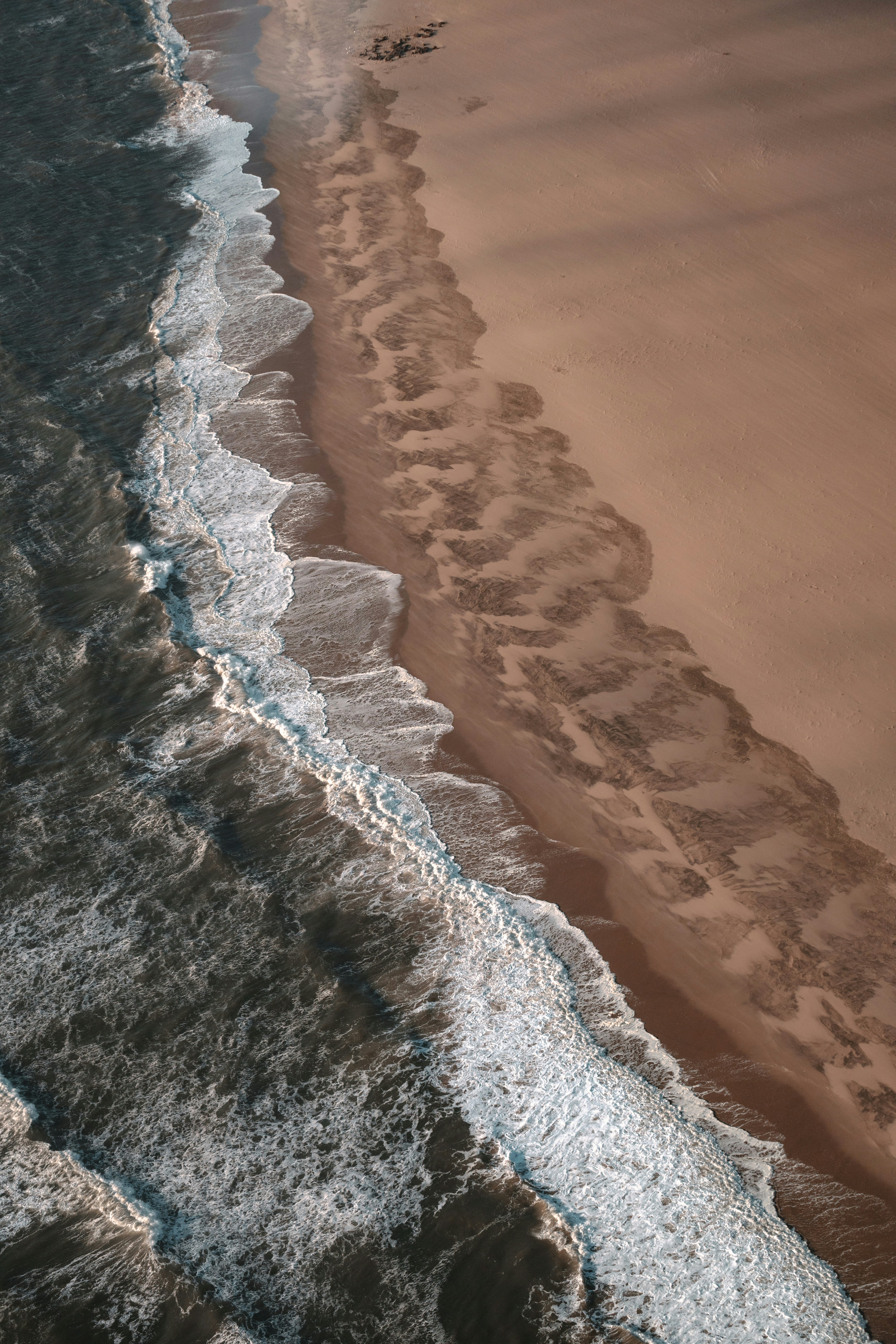 Aerial photograph of a coastal shoreline where dark ocean meets warm sand, with white foam tracing a diagonal boundary.
