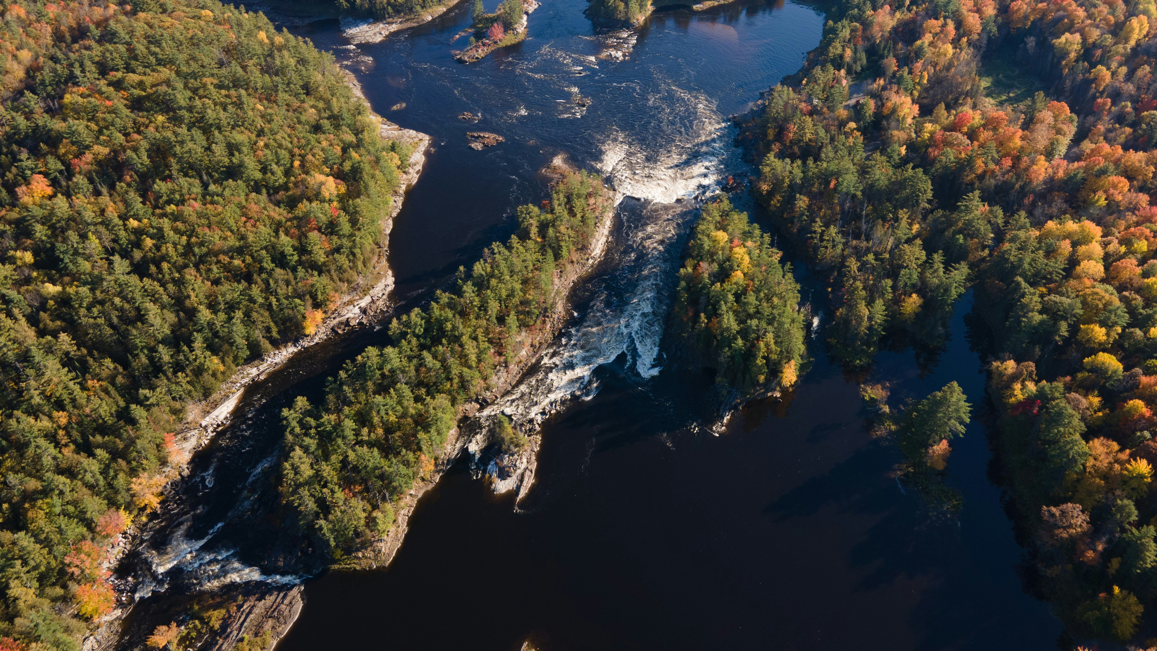 Aerial view of a winding river surrounded by vibrant autumn foliage, showcasing the interplay of water and colorful trees. Water cascades over rocks, adding dynamic movement to the serene landscape.