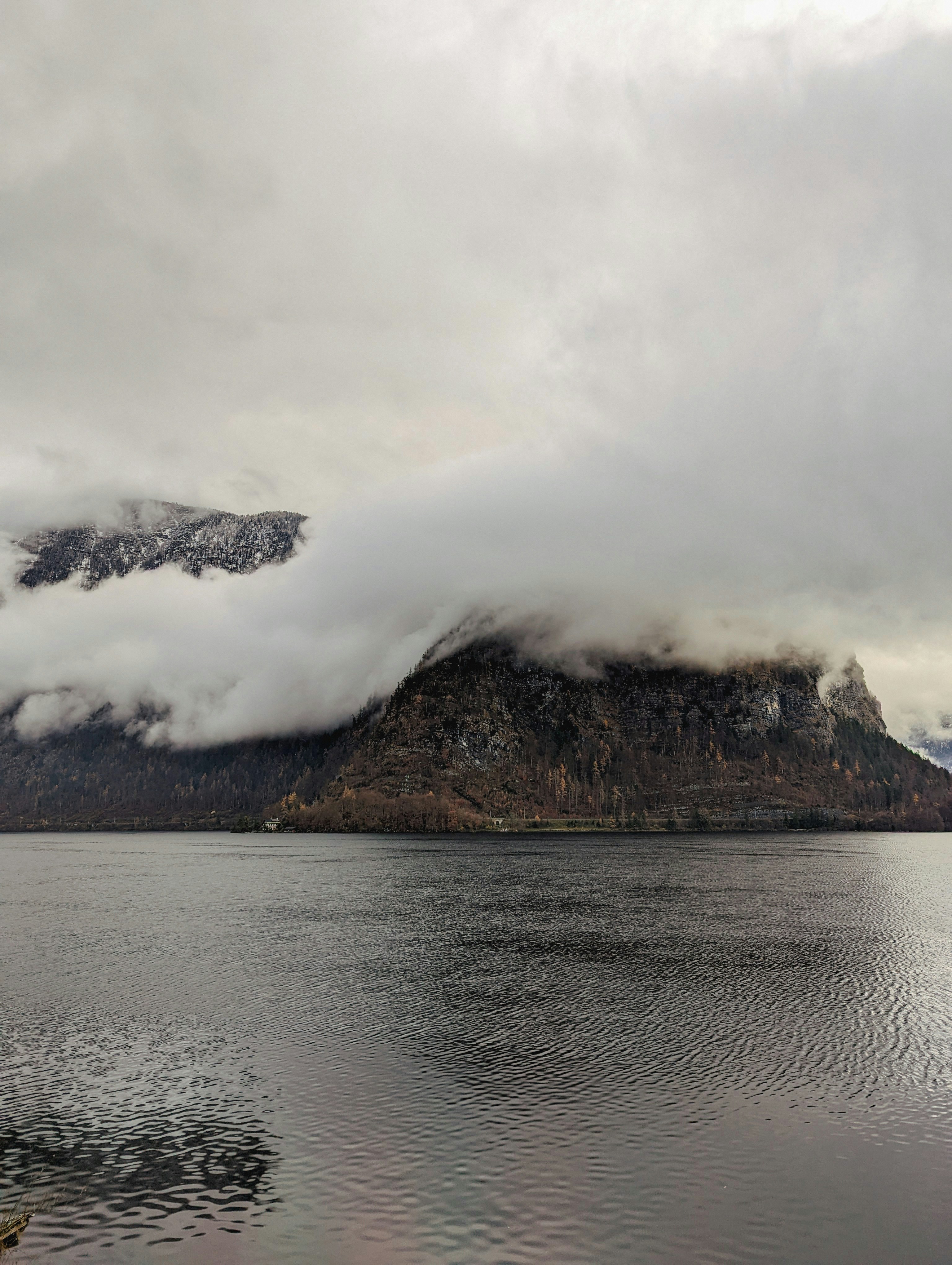 Low-hanging clouds enveloping a rugged mountain landscape beside a tranquil lake, reflecting the muted tones of the overcast sky.