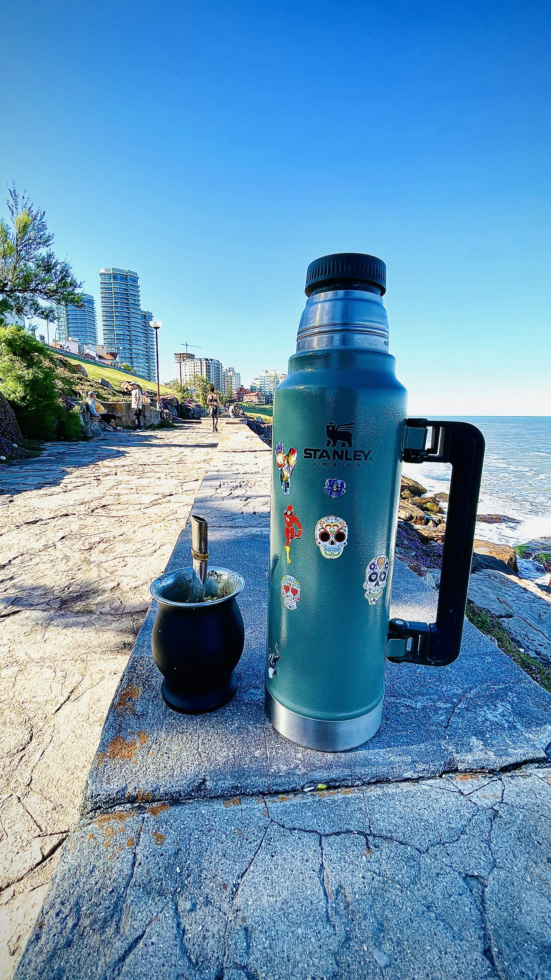 A Stanley thermos adorned with colorful stickers sits beside a traditional mate cup on a stone pathway, overlooking a serene coastal view with city buildings in the background.