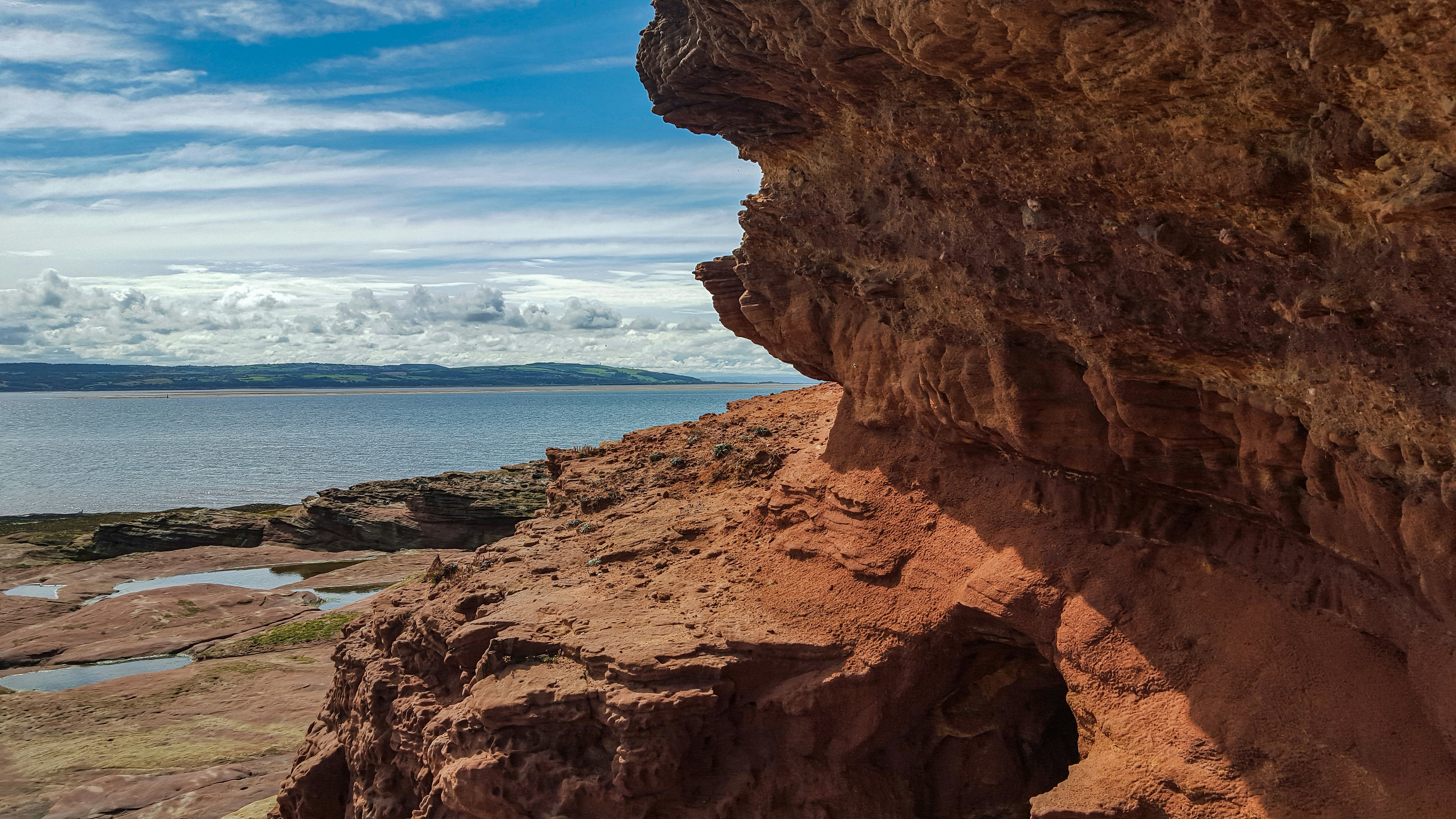 Sunlit red rock formation by the shoreline with calm water in the background; a coastal cliff edge framing the scene. The image captures texture and color of the rock and the tranquil seascape.