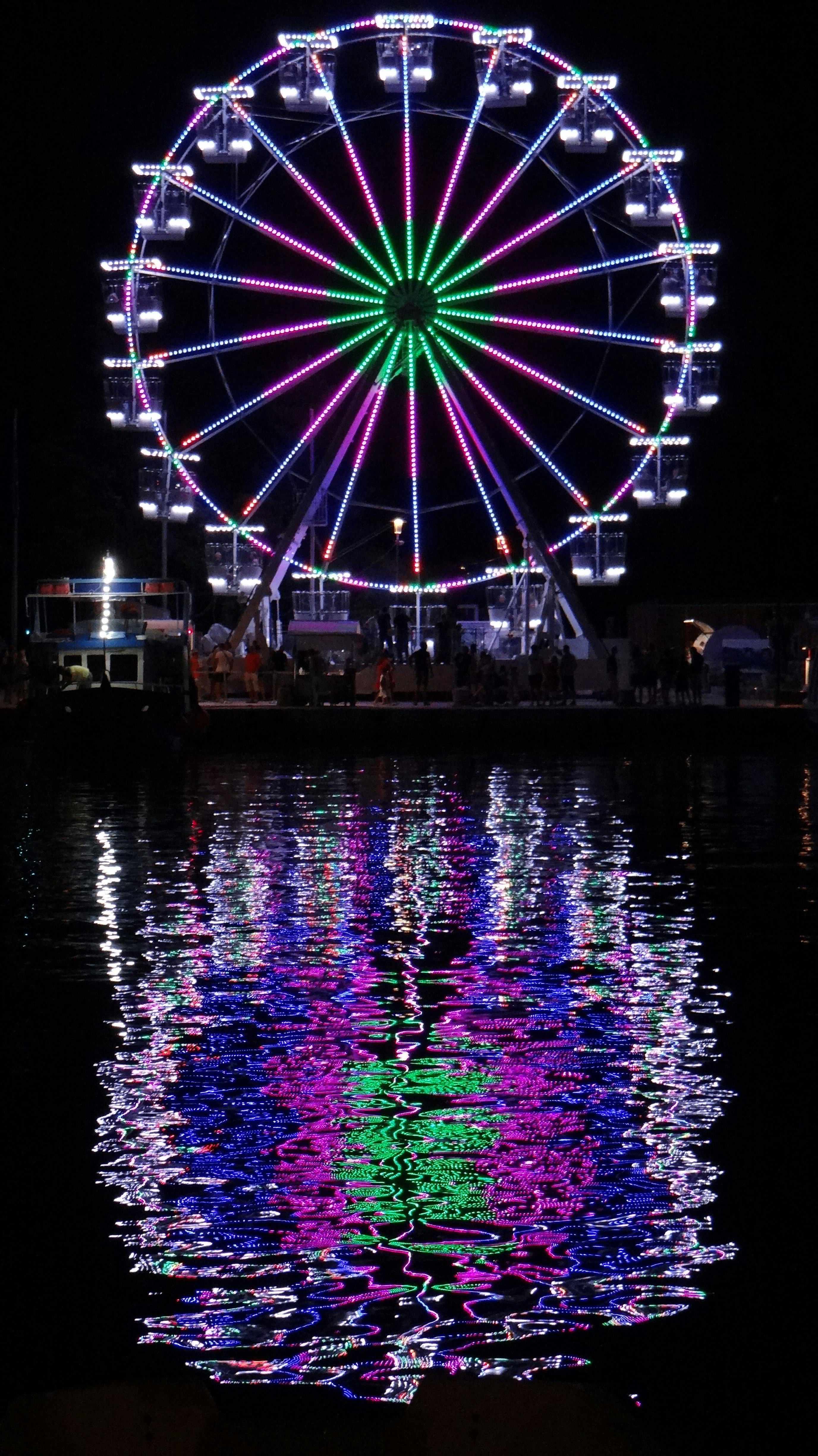 A vibrant Ferris wheel illuminated with colorful lights, reflecting beautifully on the water's surface at night.