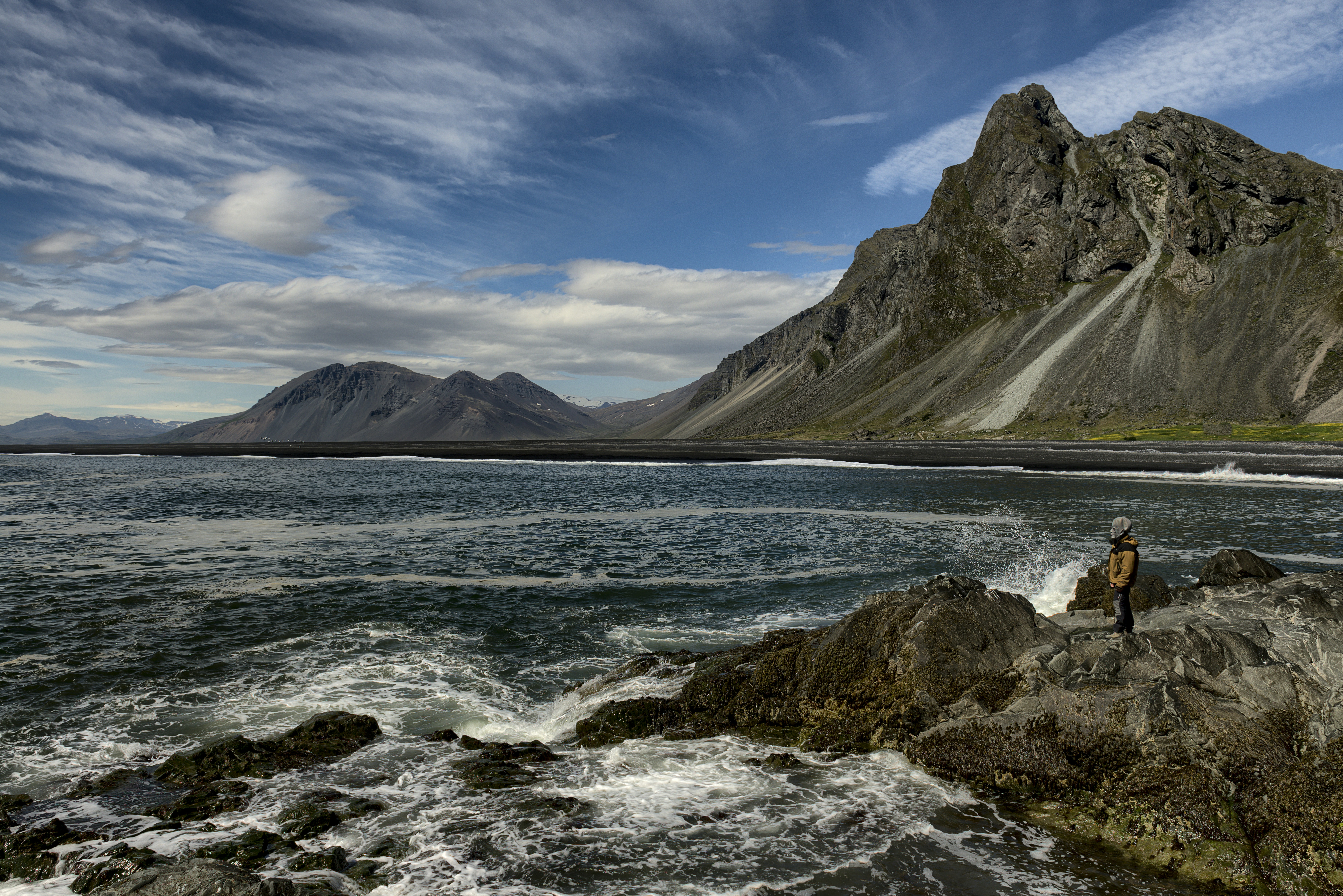 Rugged Coastal Portrait: A Seeker on Jagged Rocks with Alpine Backdrop