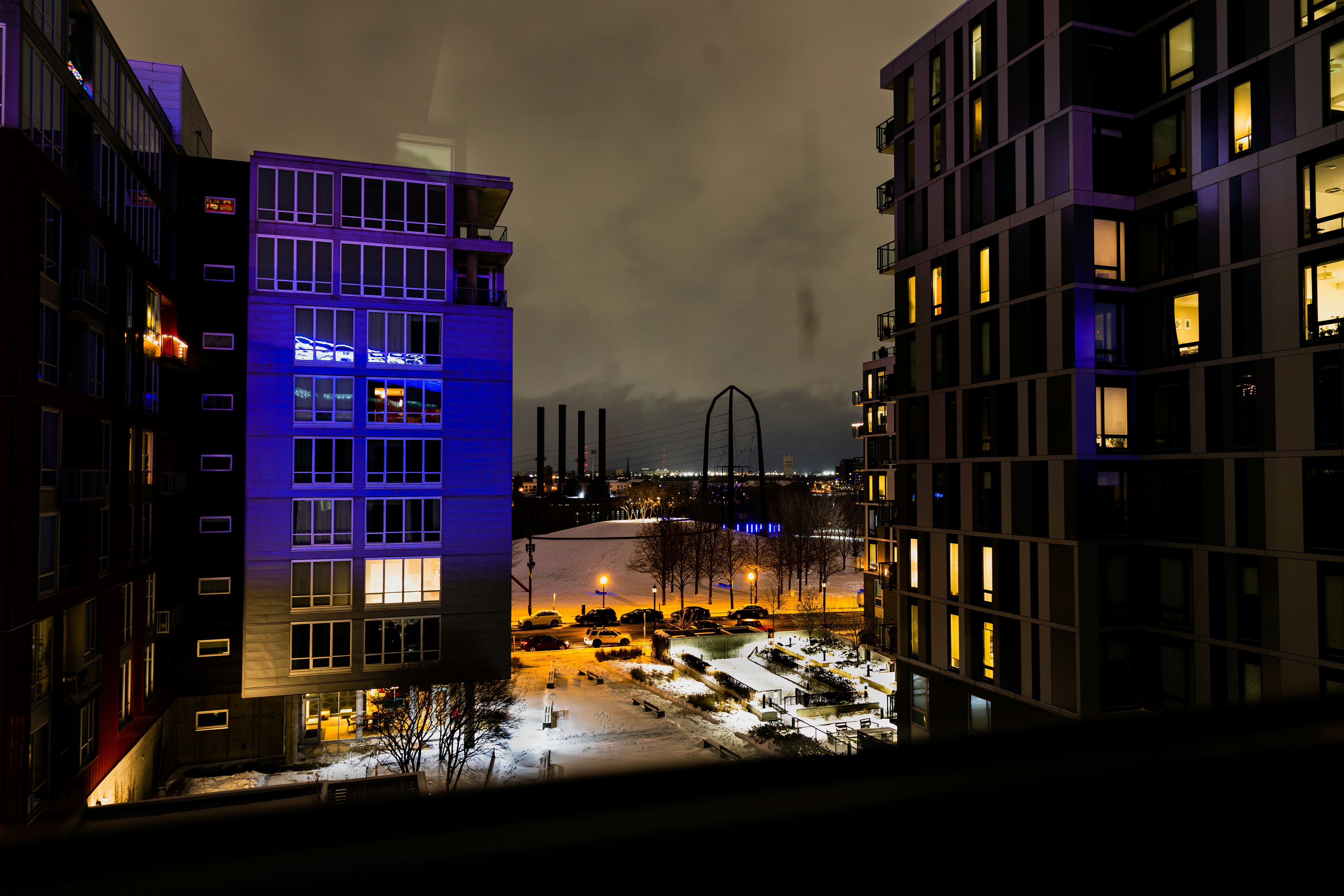 Cityscape view between two buildings at night, with illuminated windows and industrial structures in the distance.