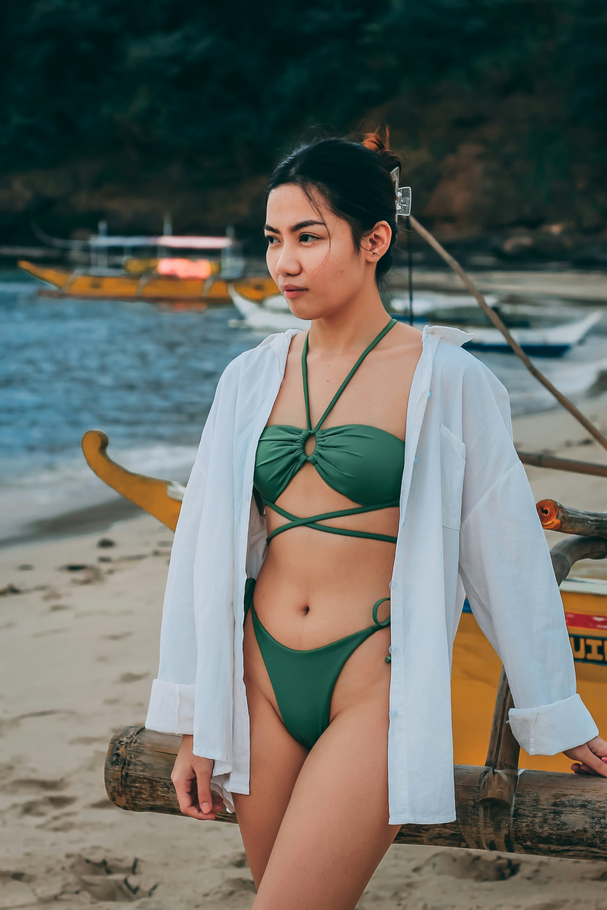 Woman in green swimsuit and white shirt standing on a sandy beach with boats in the background.
