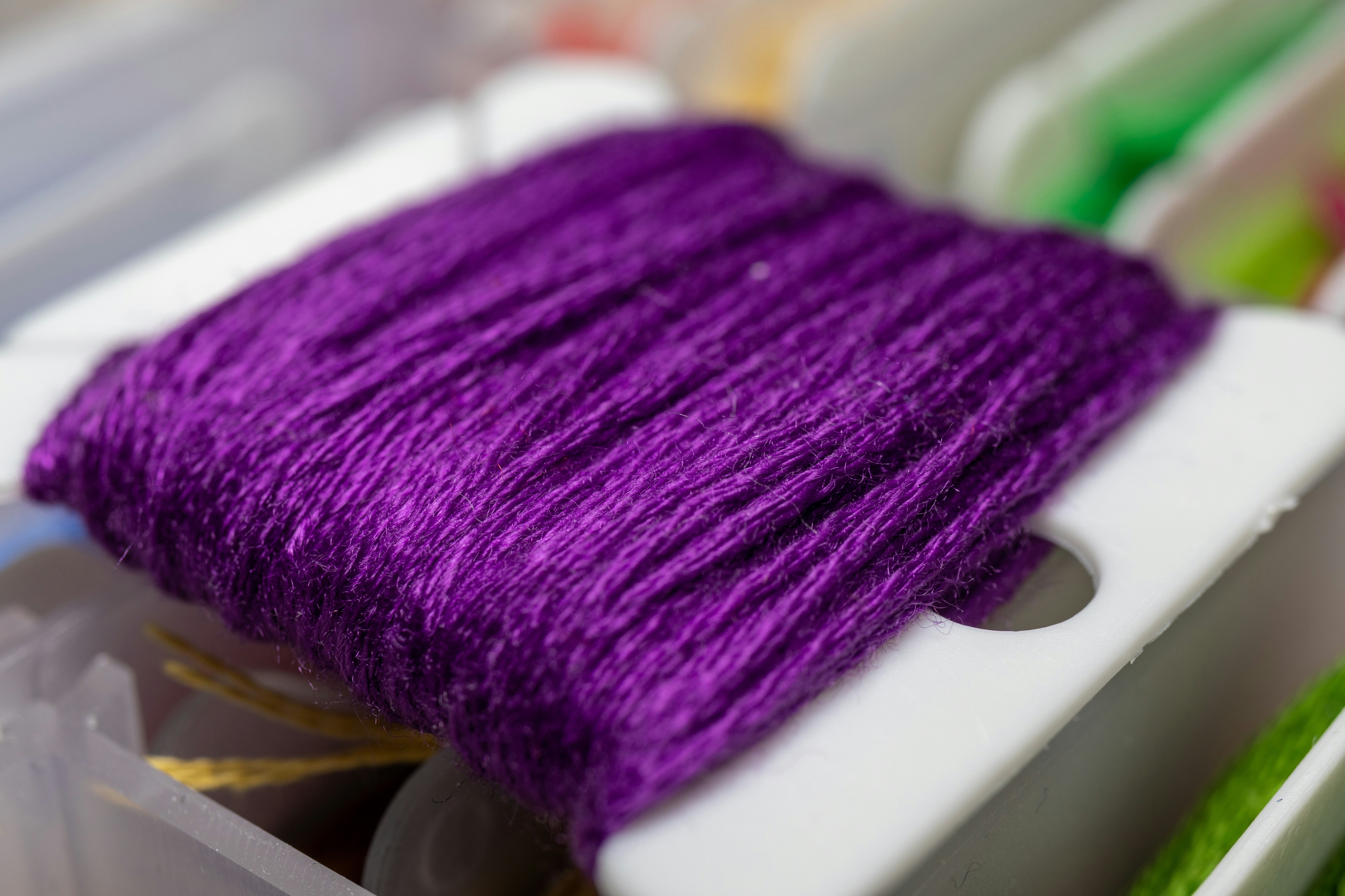 Close-up of vibrant purple thread wound around a white bobbin, surrounded by blurred hues of other colors.