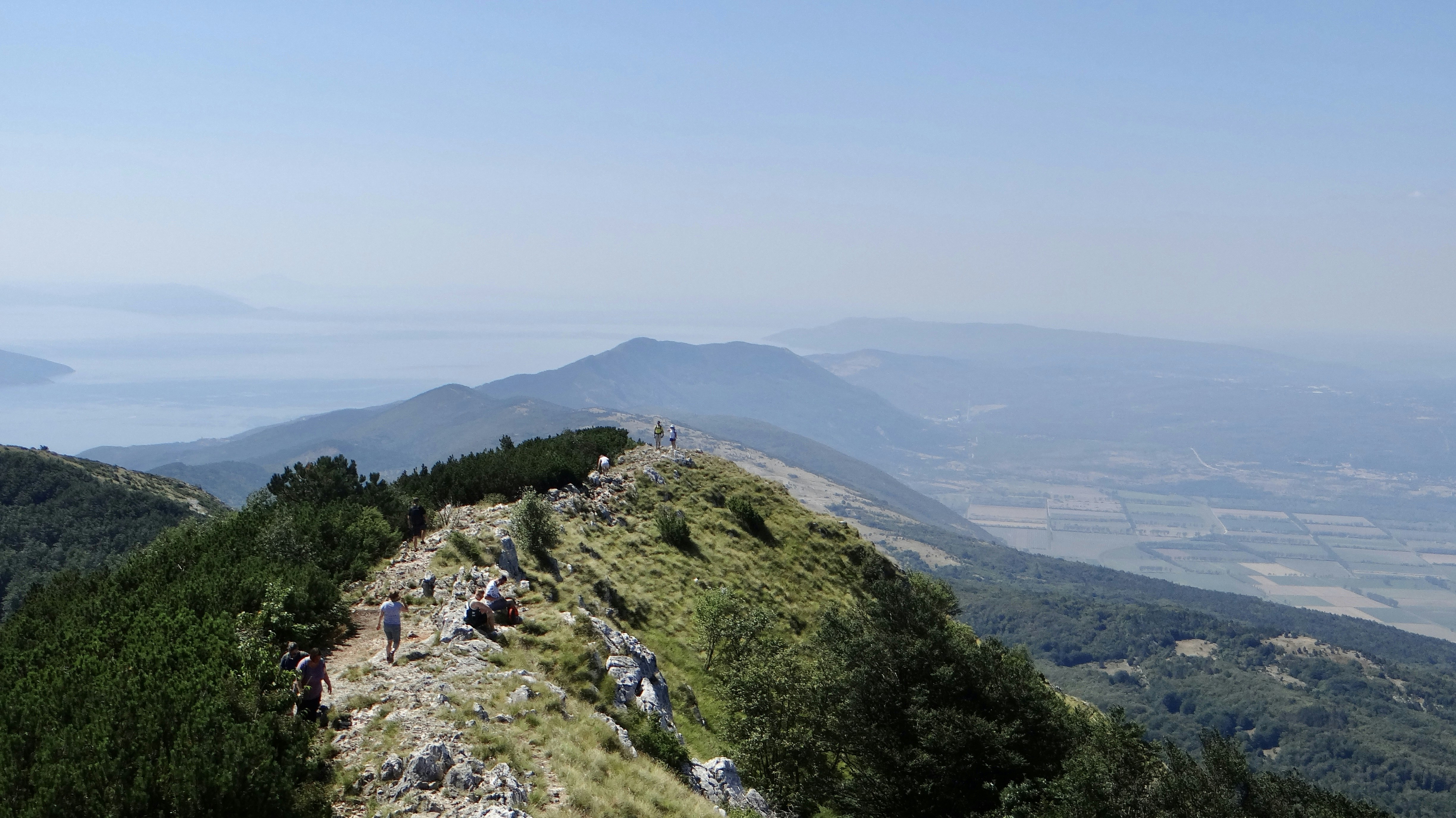 Hikers traverse a rocky ridge overlooking a vast landscape of mountains and valleys under a clear blue sky.