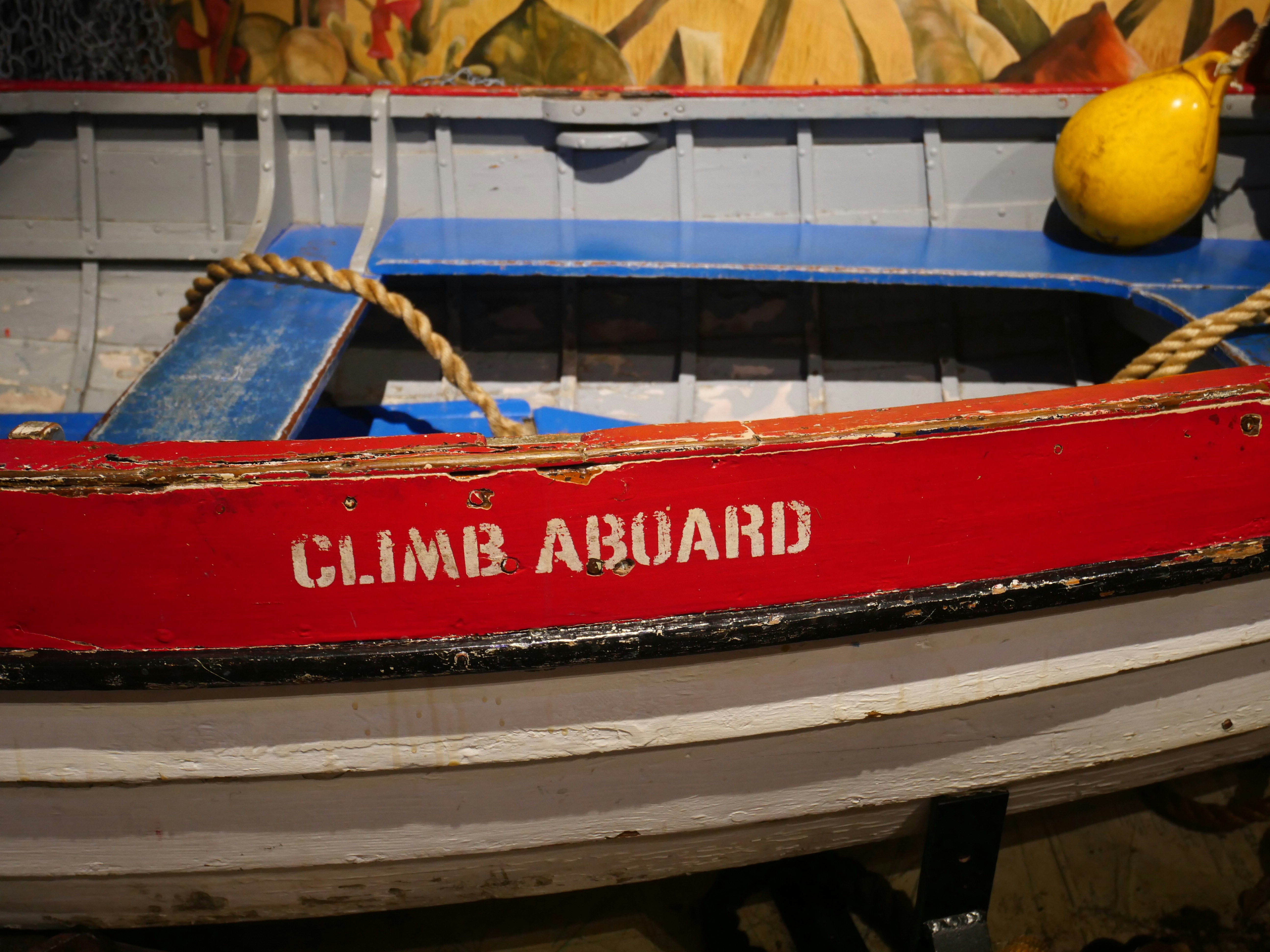 Close-up of a red boat's bow featuring the phrase 'CLIMB ABOARD,' complemented by a yellow buoy and blue accents. The background hints at a vibrant maritime theme.