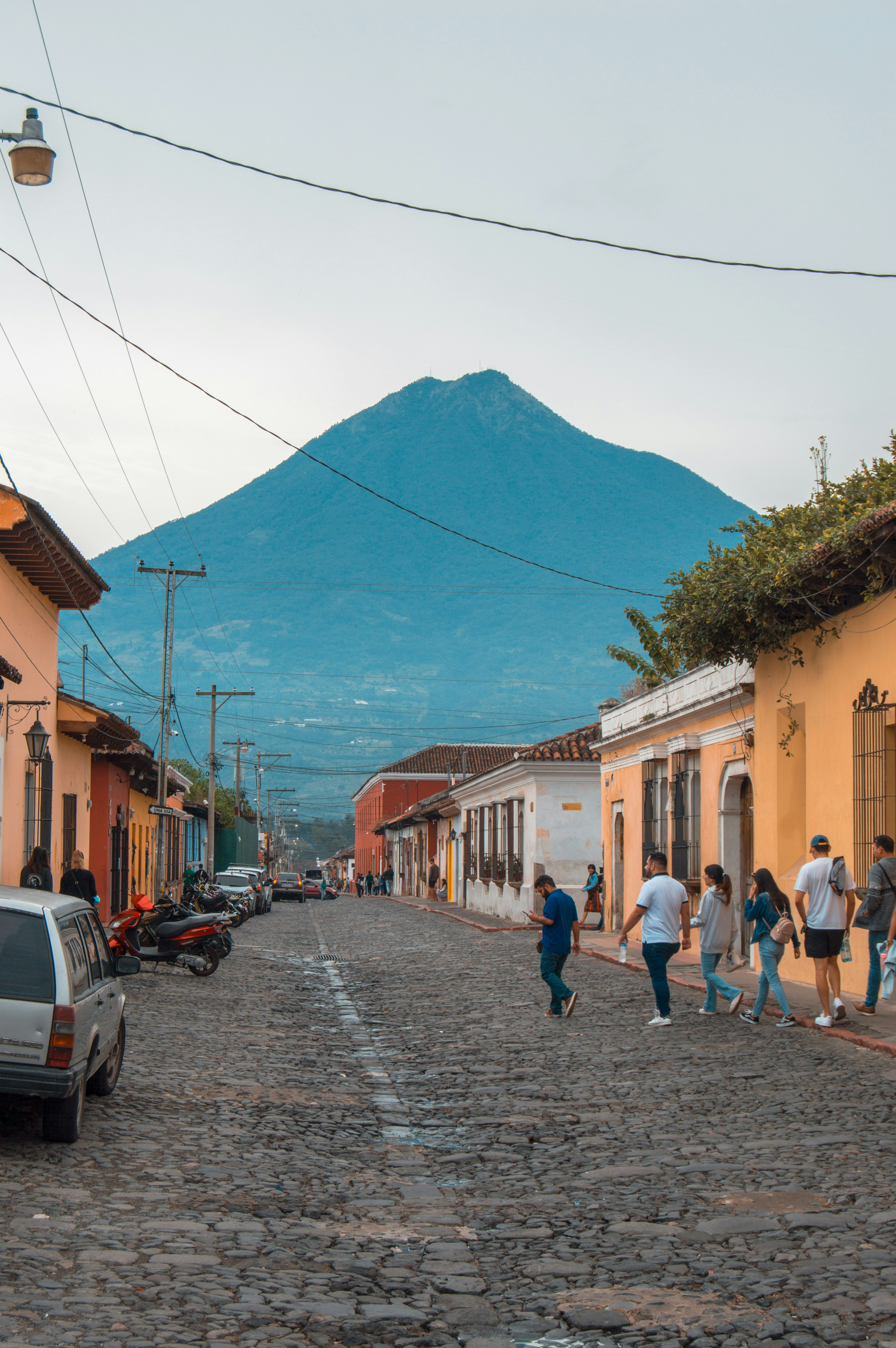 Cobblestone street in Antigua, Guatemala, bustling with pedestrians and framed by colonial architecture, with a volcano looming in the background.