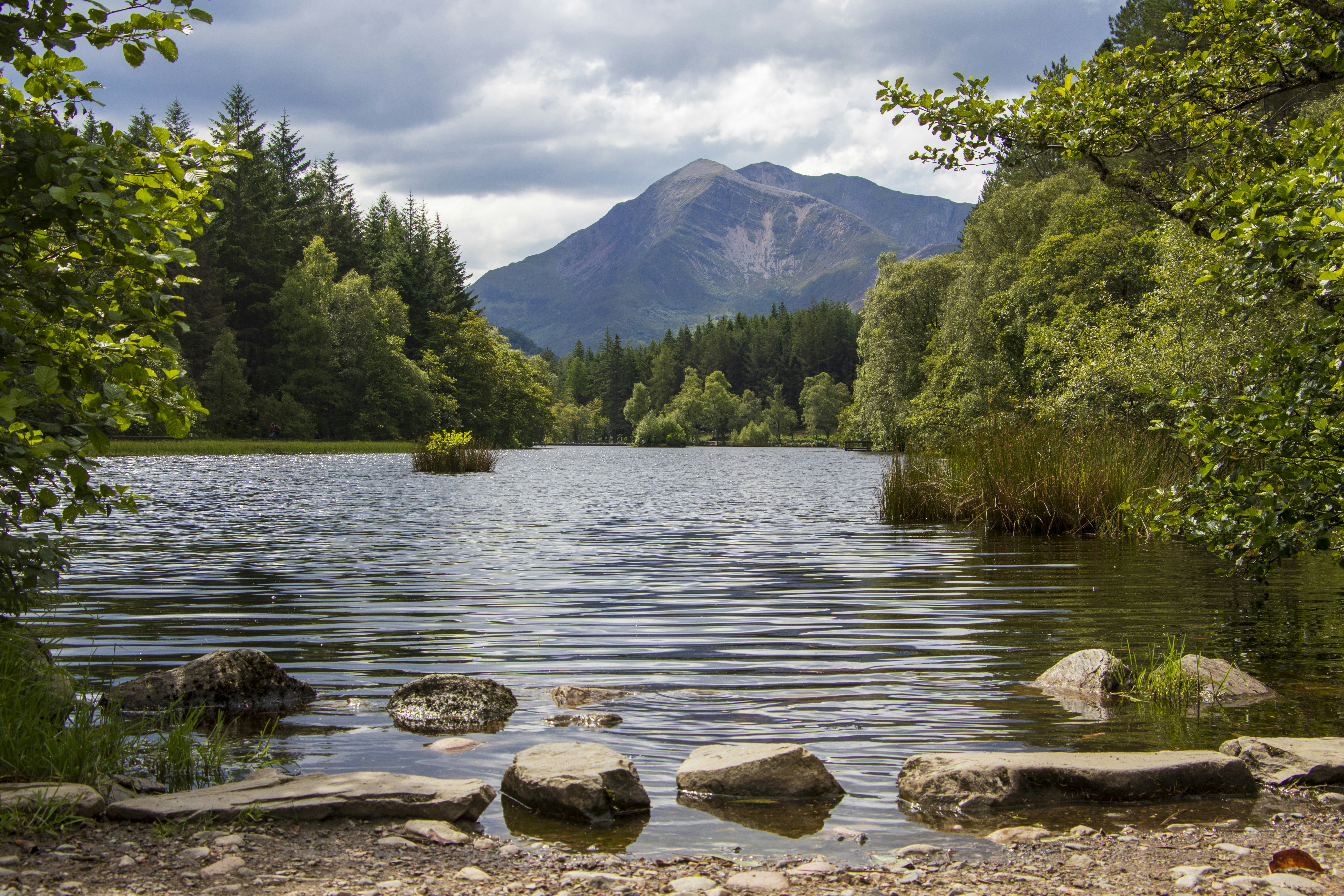 Tranquil lake surrounded by lush greenery and distant mountains under a cloudy sky.