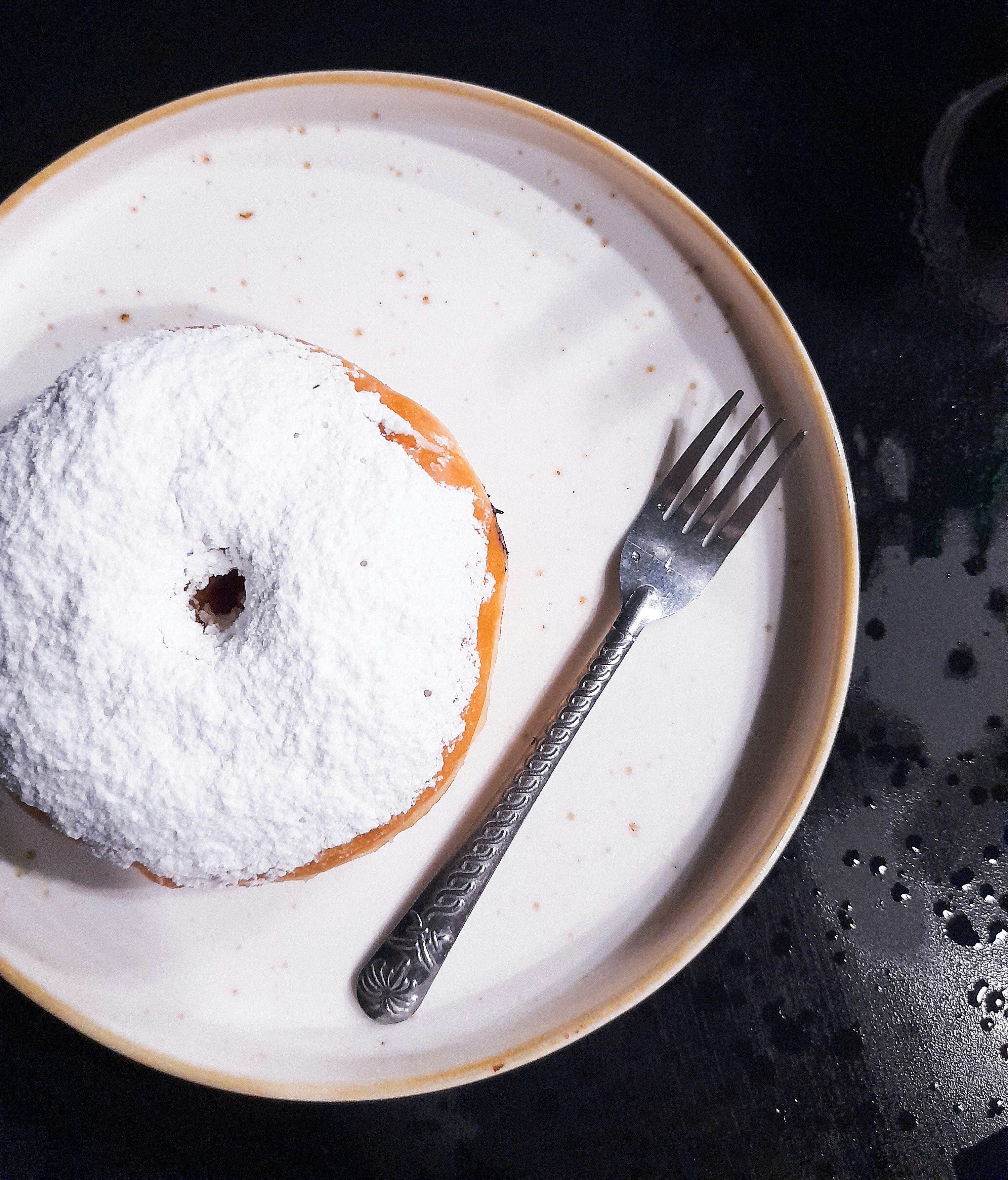 A fluffy donut dusted with powdered sugar sits on a speckled plate beside a vintage fork, showcasing a tempting treat ready to be enjoyed.
