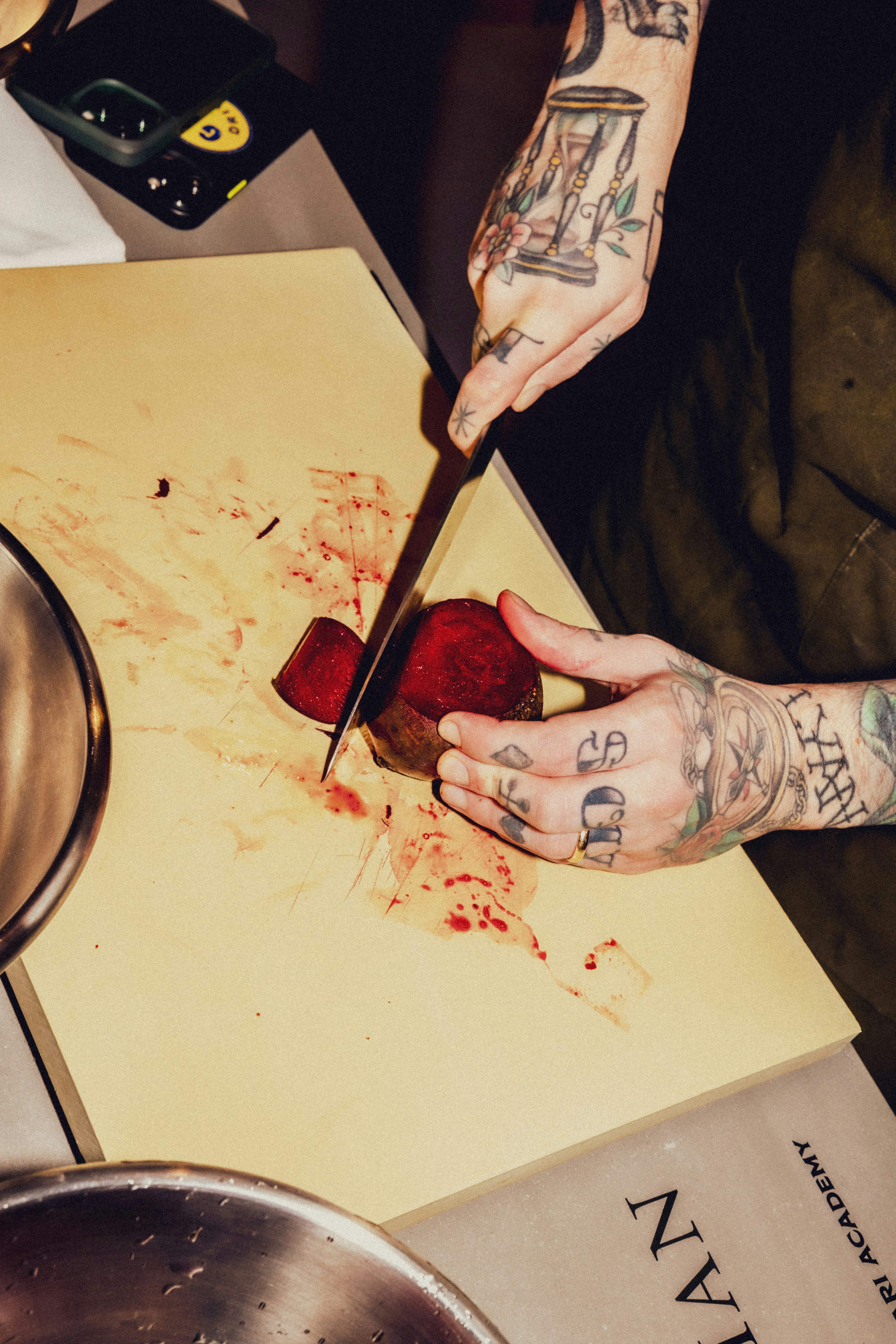 A chef skillfully slicing beets on a yellow cutting board, showcasing intricate tattoos and vibrant colors in a kitchen setting.