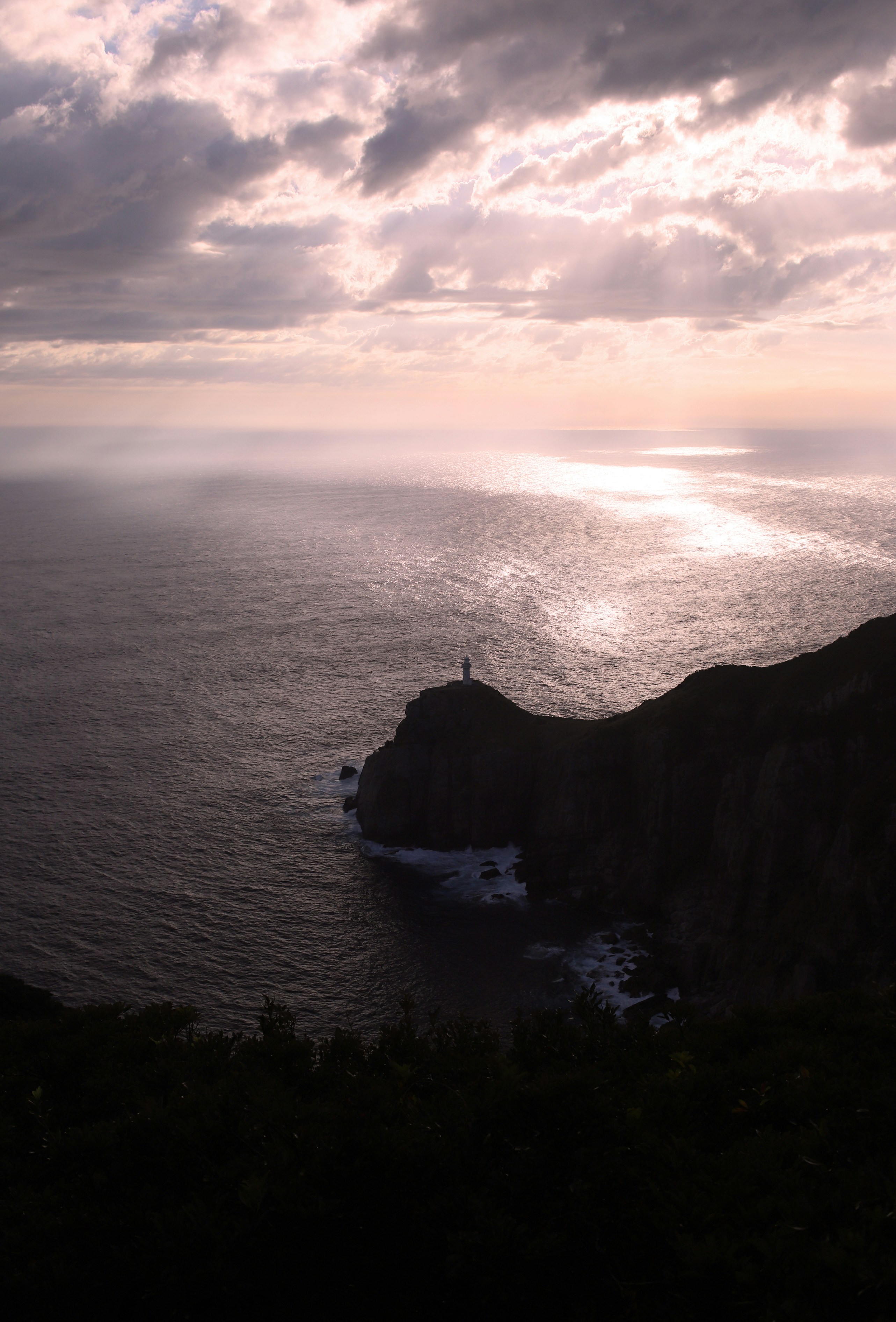 A lighthouse stands tall on a rugged cliff, surrounded by the vast expanse of the ocean under a dramatic sky. The shimmering water reflects the soft light of the setting sun.