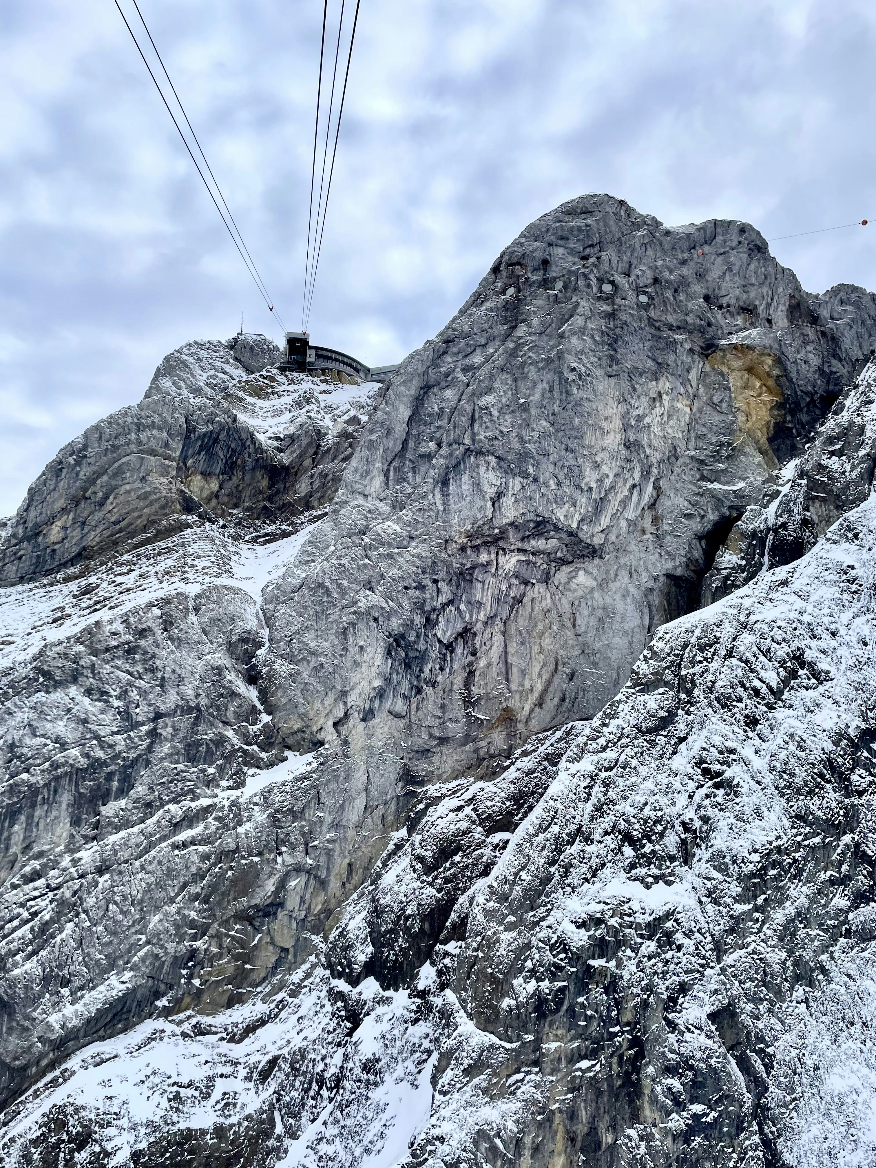 Snow-dusted jagged cliffs frame a distant ropeway station perched near a rocky summit.
