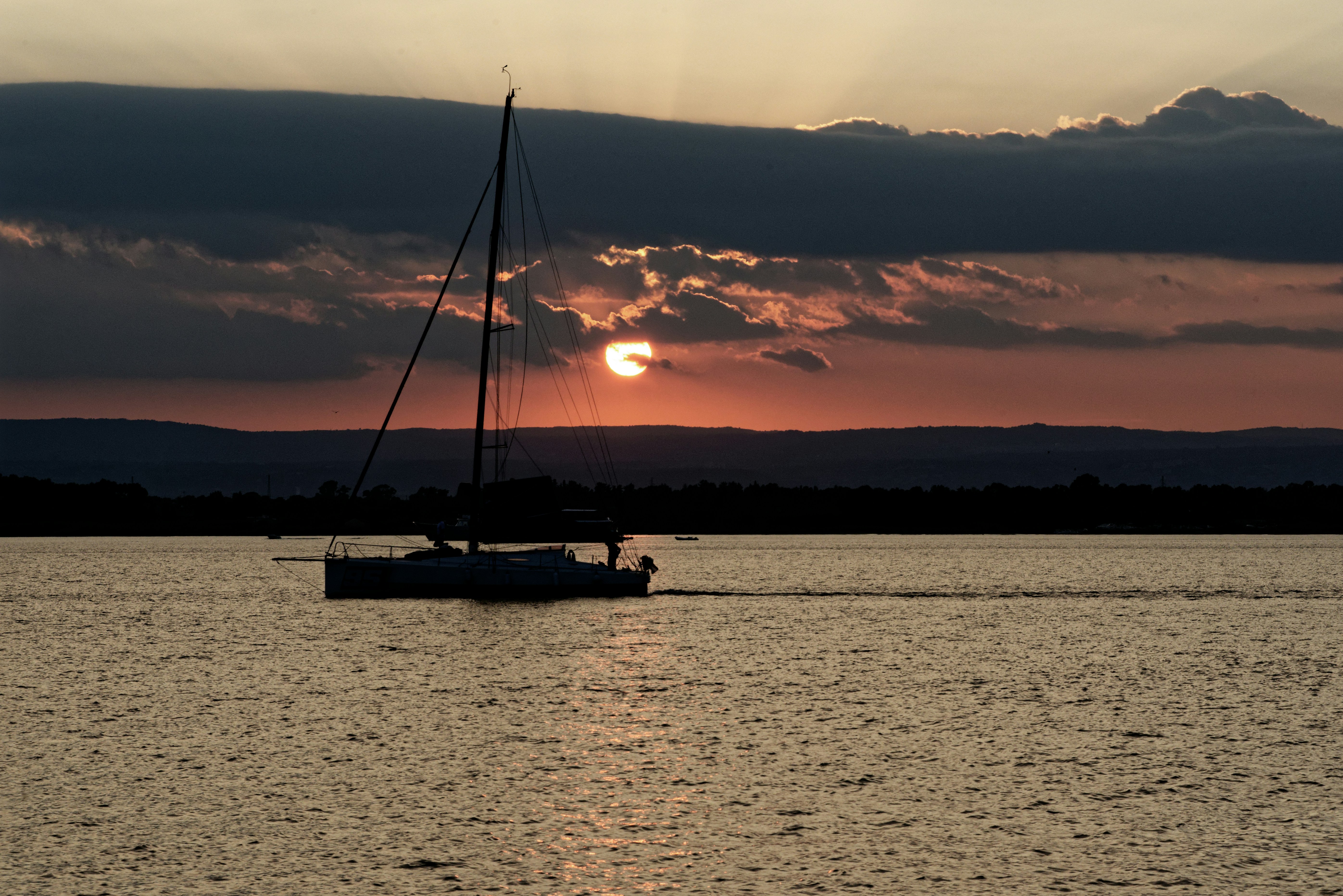 Silhouette of a sailboat gliding across calm waters at sunset with a dramatic sky overhead.