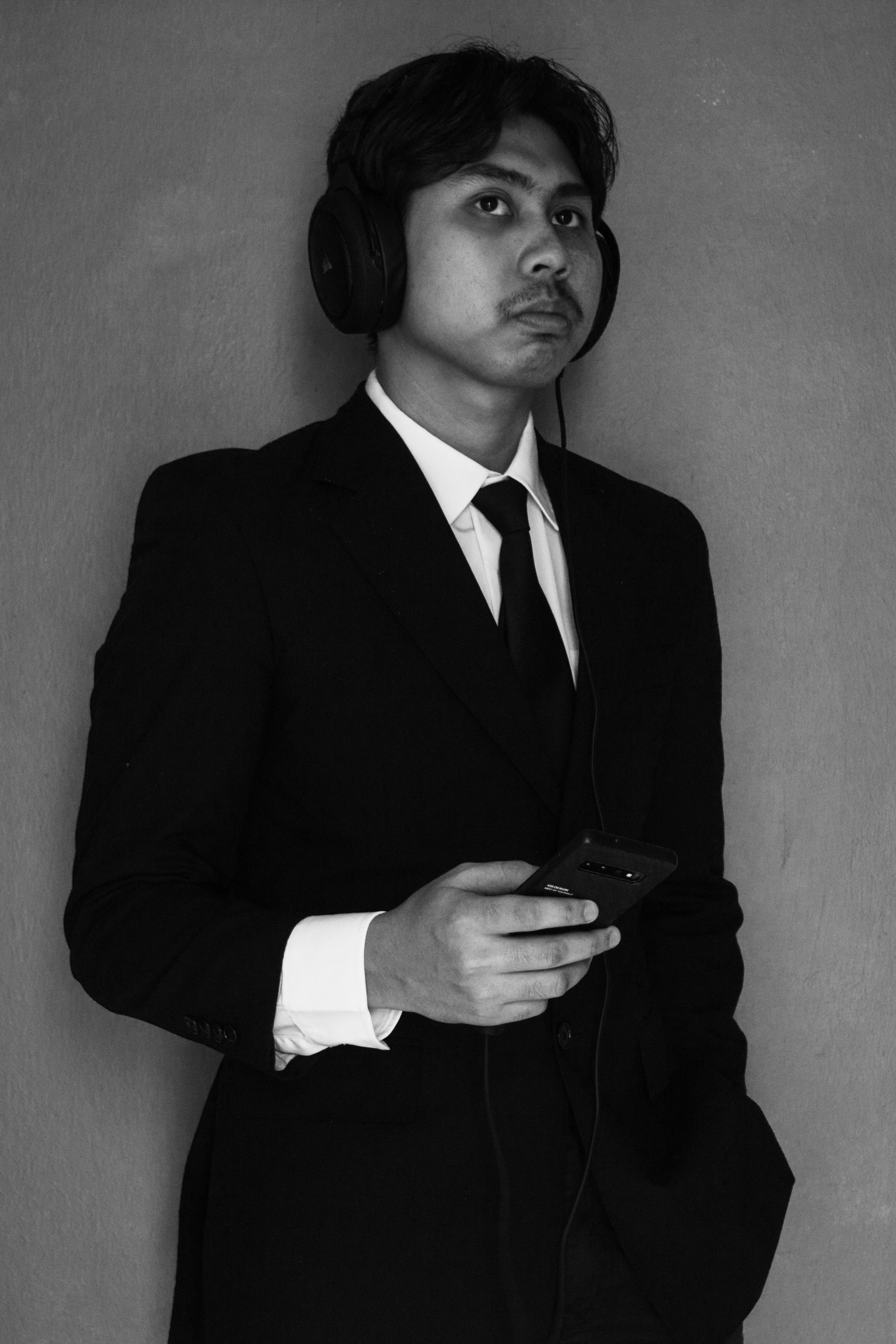 A man in a suit, wearing headphones, stands against a textured background while holding a smartphone. The monochrome tones emphasize the mood of contemplation.