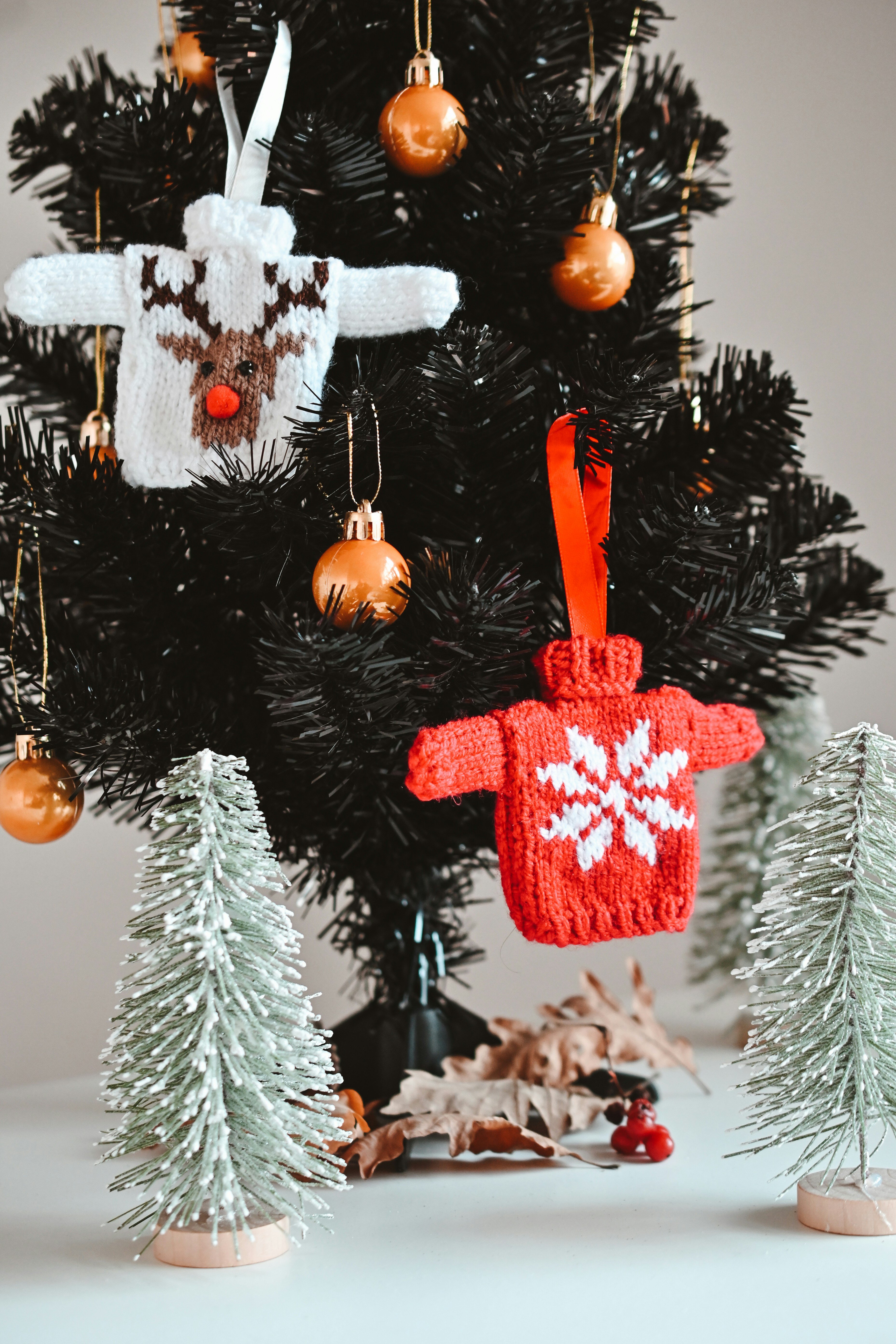 Close-up of a small Christmas tree featuring knitted ornaments, including a red snowflake sweater ornament in the foreground and a white reindeer ornament nearby, amid gold baubles.