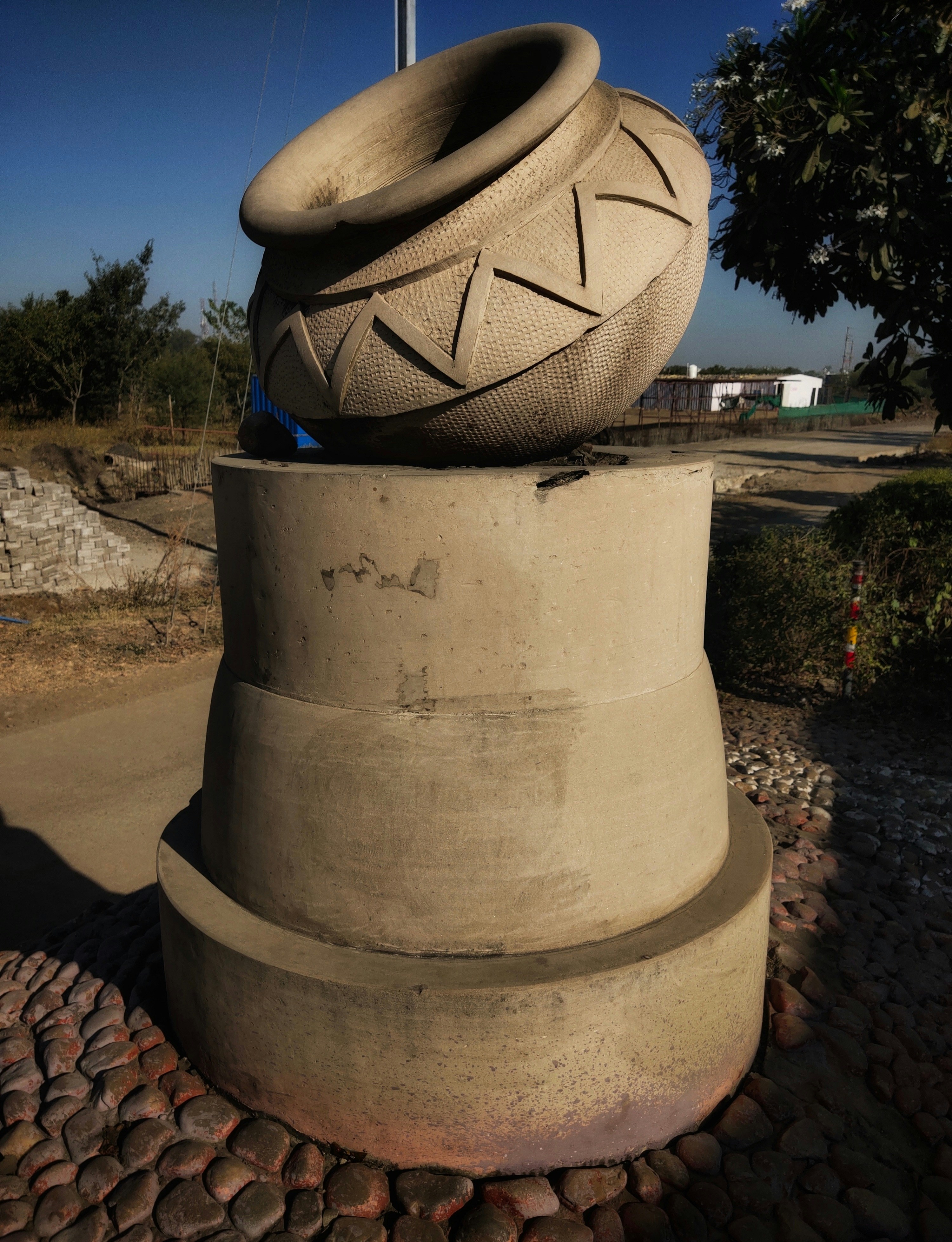 A textured clay pot elegantly balanced atop a cylindrical base, showcasing craftsmanship against a serene outdoor backdrop.