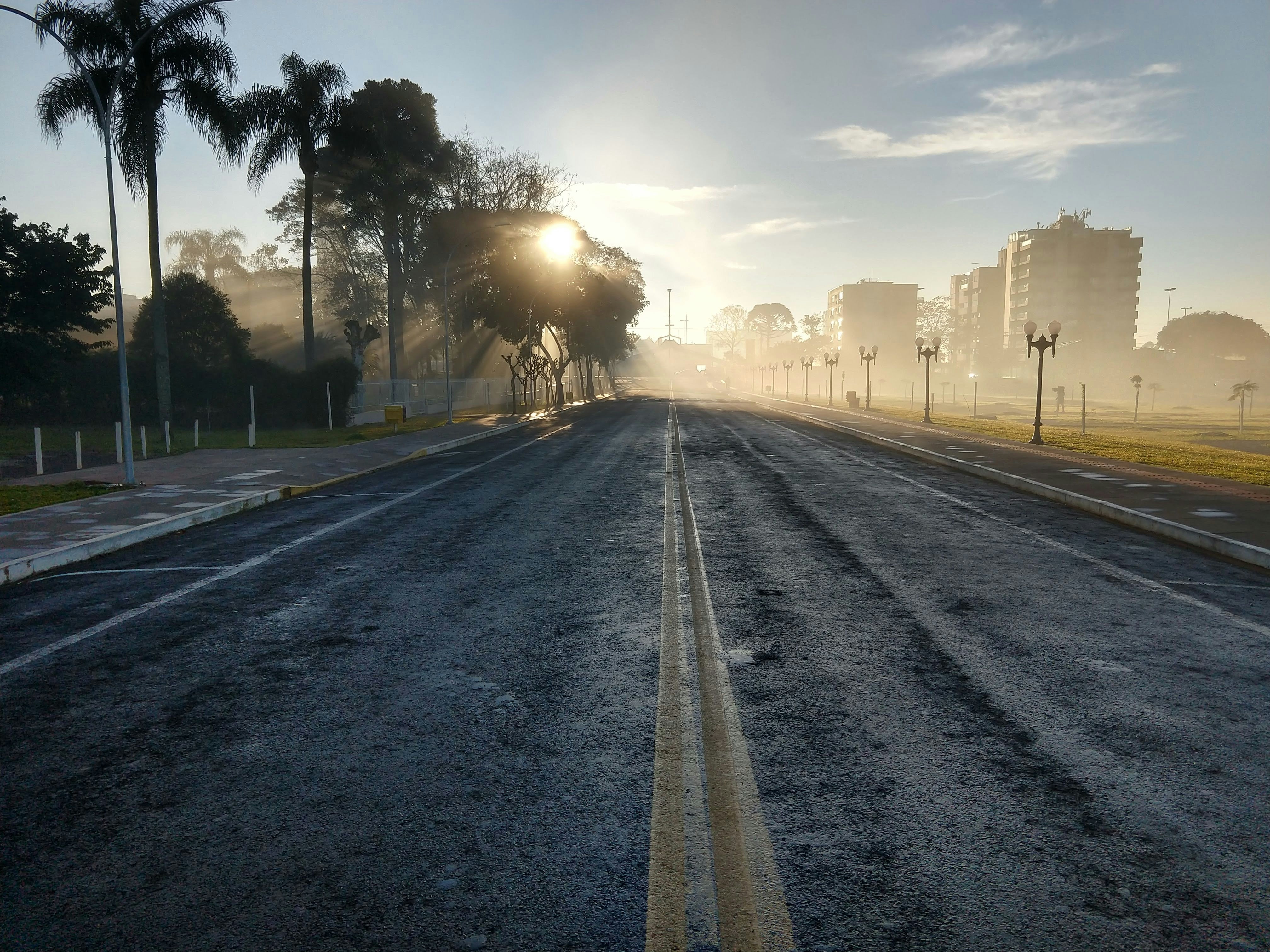 Sunlight breaks through mist over an empty road lined with trees and buildings.