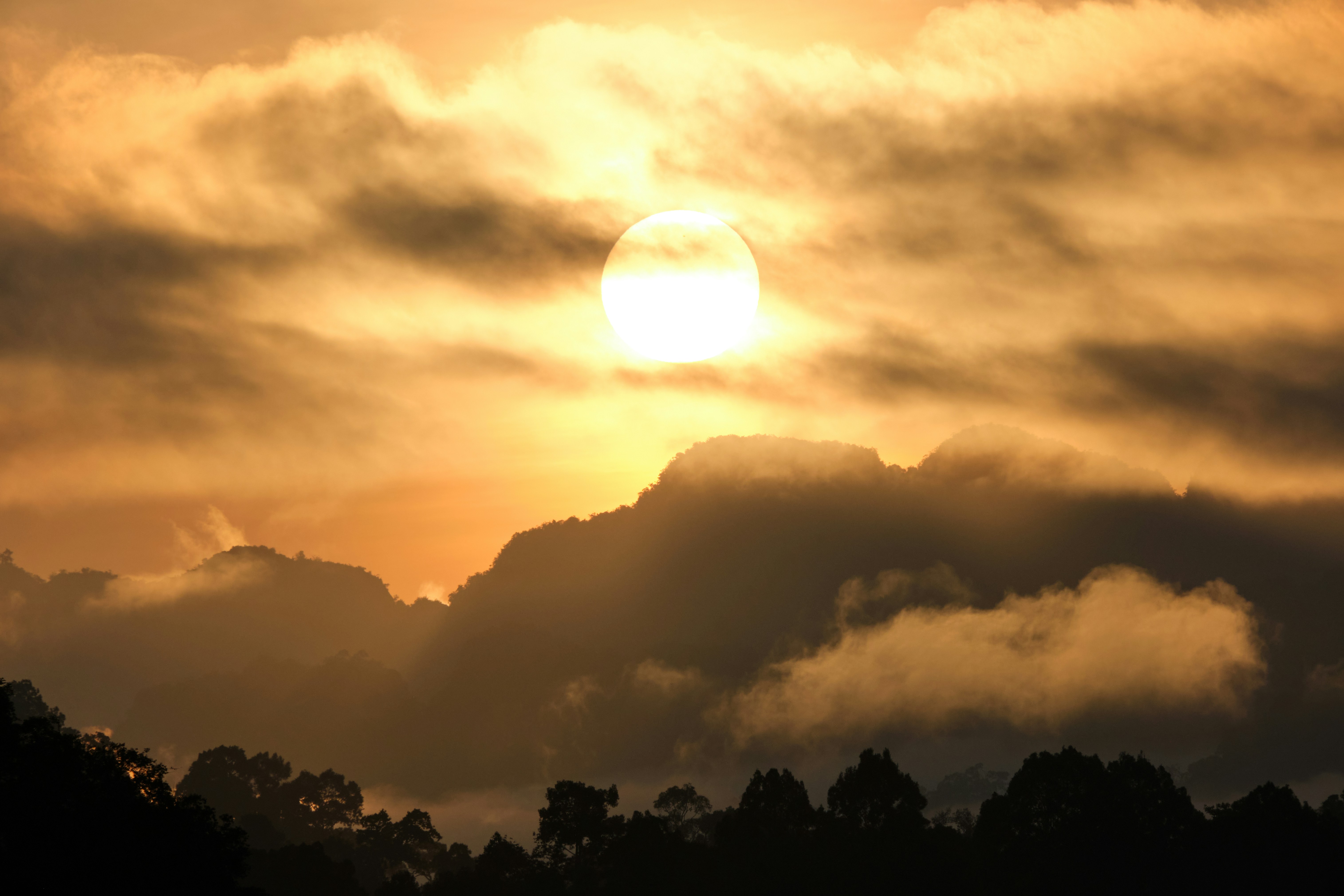 Sun setting behind clouds with silhouetted mountains and trees in the foreground.