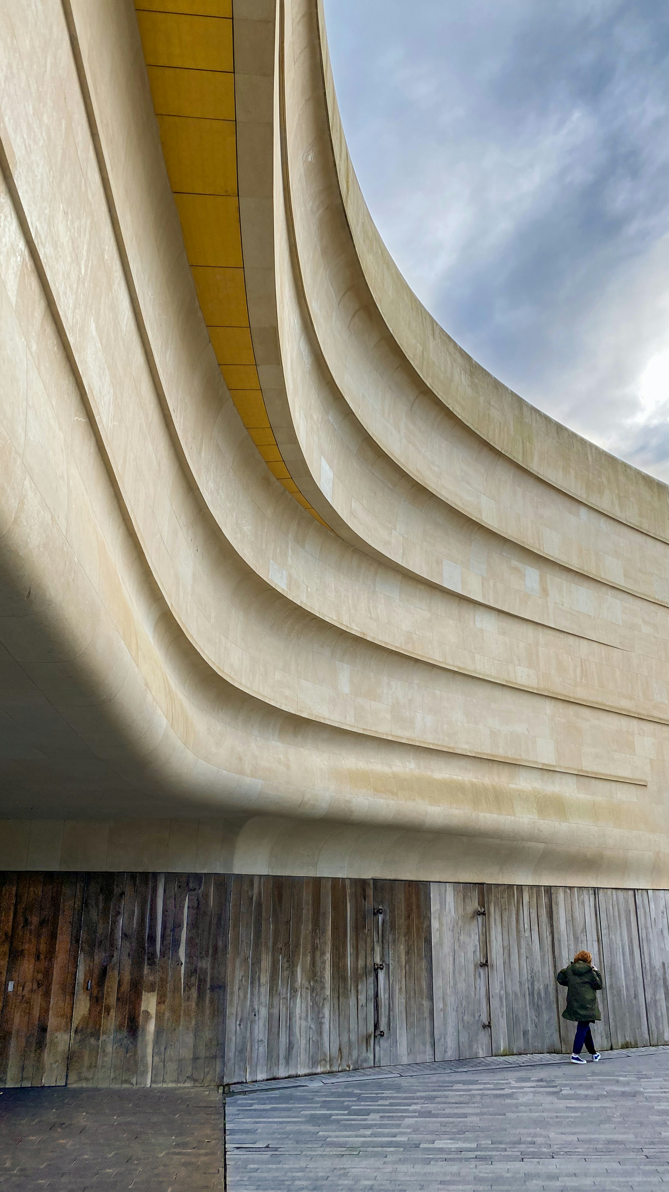Architectural abstraction featuring smooth, flowing lines of a modern building, with a solitary figure walking beneath. The contrast between the curves and the wooden elements adds depth.