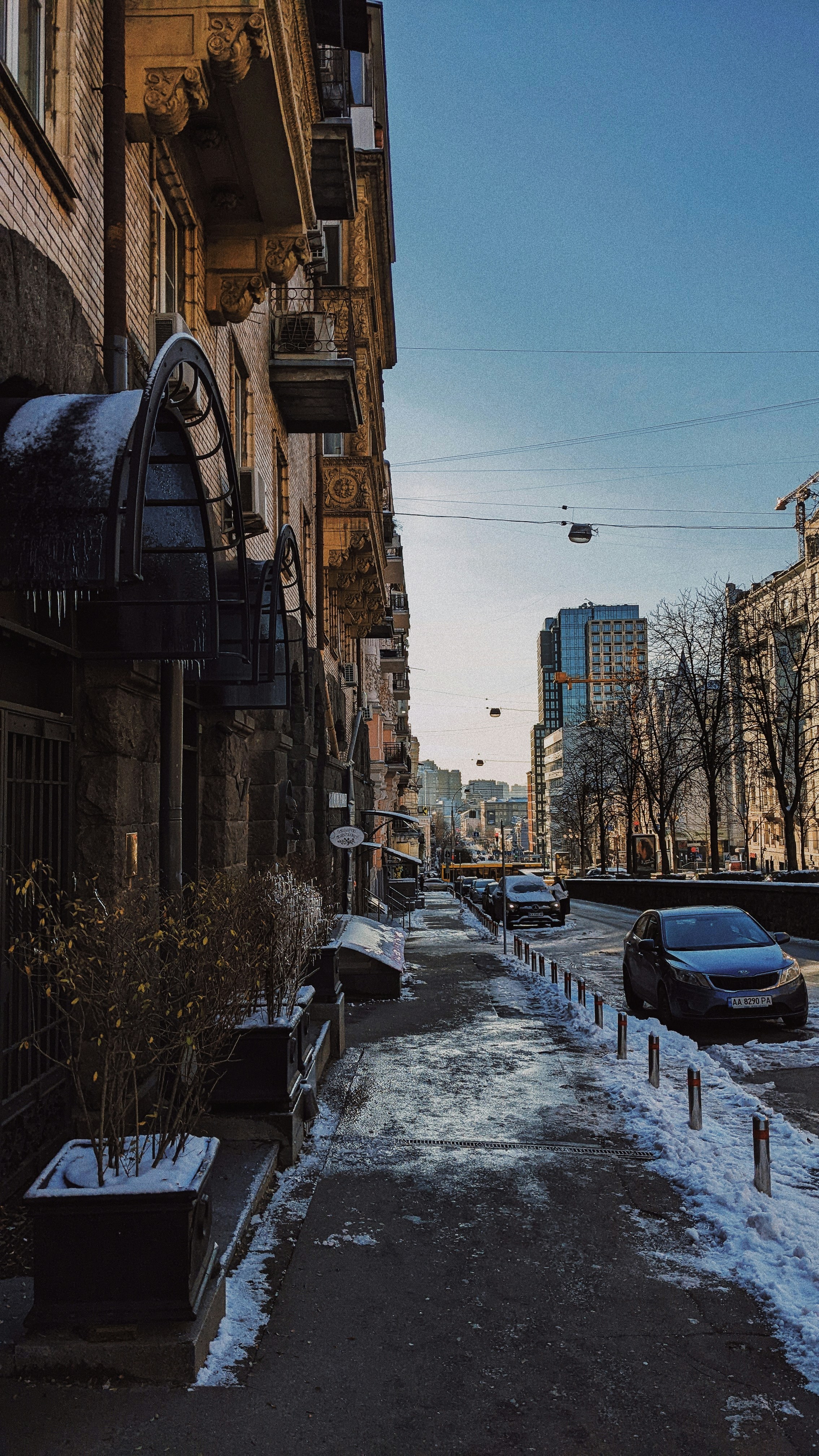 Snow-dusted city street with ornate façades and parked cars along the curb, receding toward a pale blue sky.