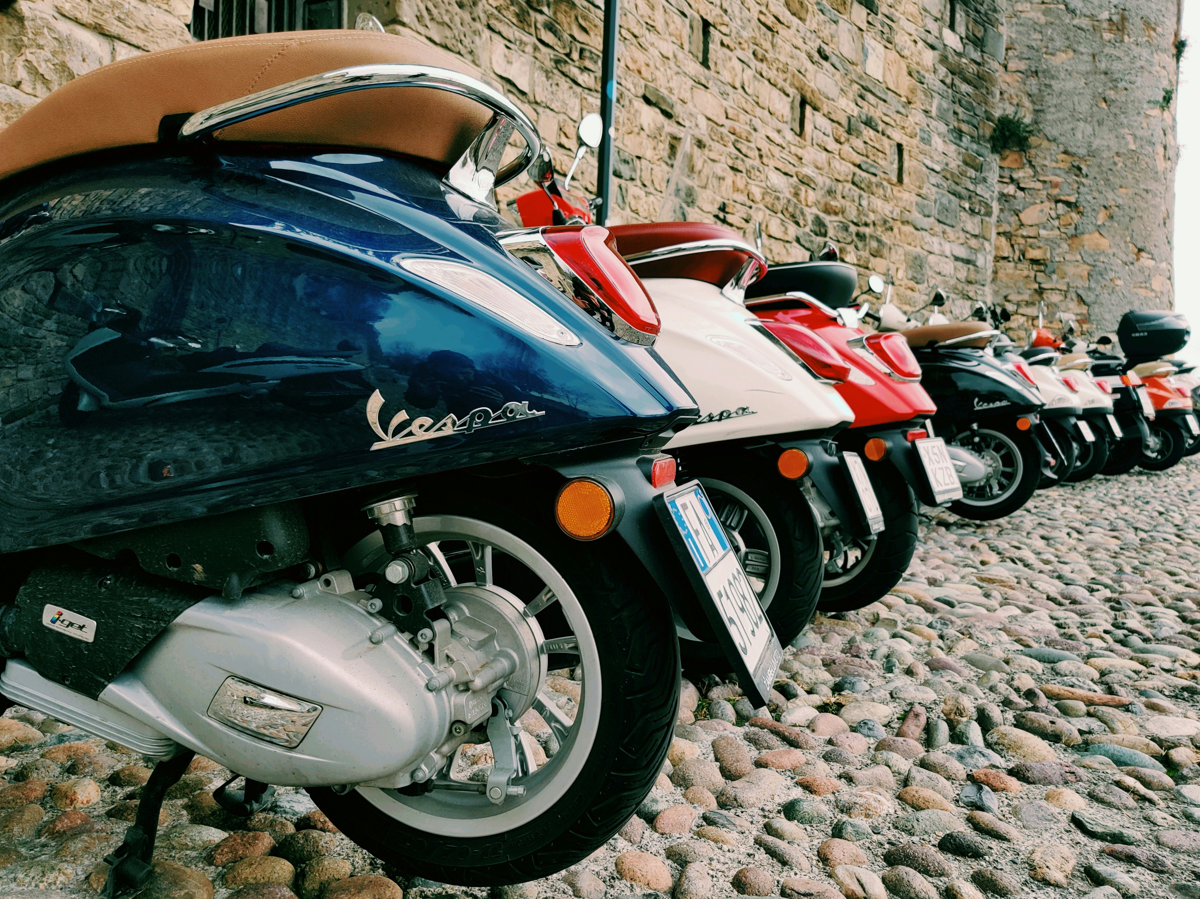 Row of colorful scooters parked along a pebble street beside a weathered stone wall. A blue Vespa dominates the foreground in this photograph.