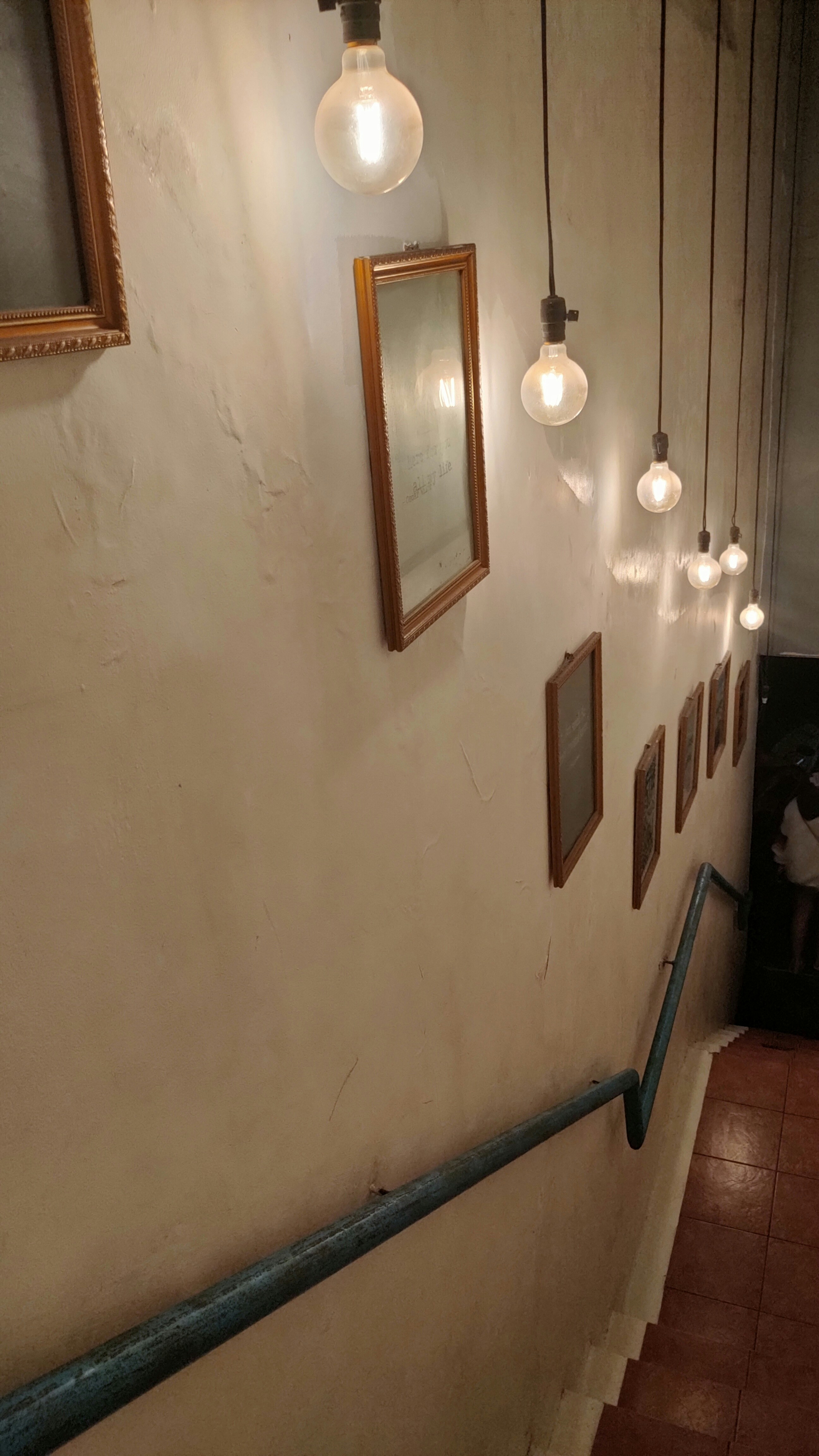 Warm light bulbs illuminate a staircase lined with framed artworks, guiding the viewer's gaze downward. The textured wall adds depth to the scene.