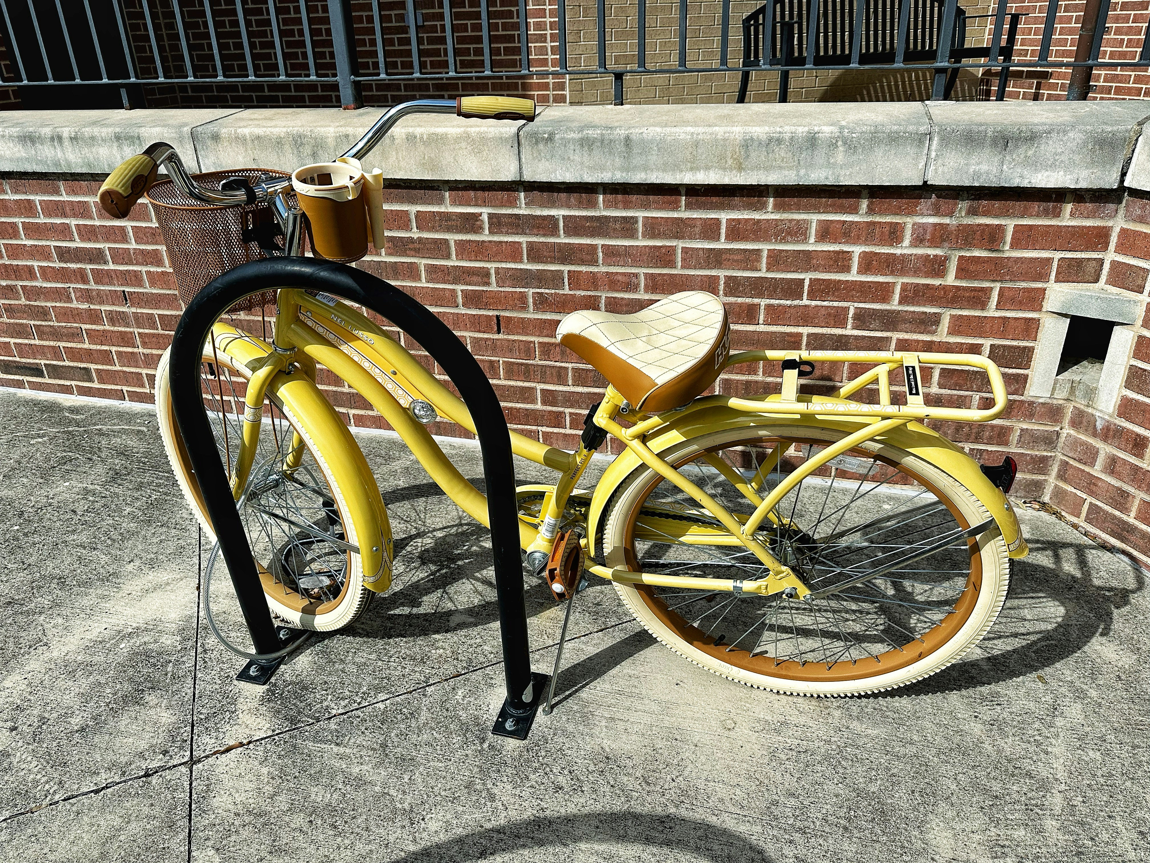 Sunlit Yellow Cruiser Locked to a Black Metal Rack on a Concrete Sidewalk