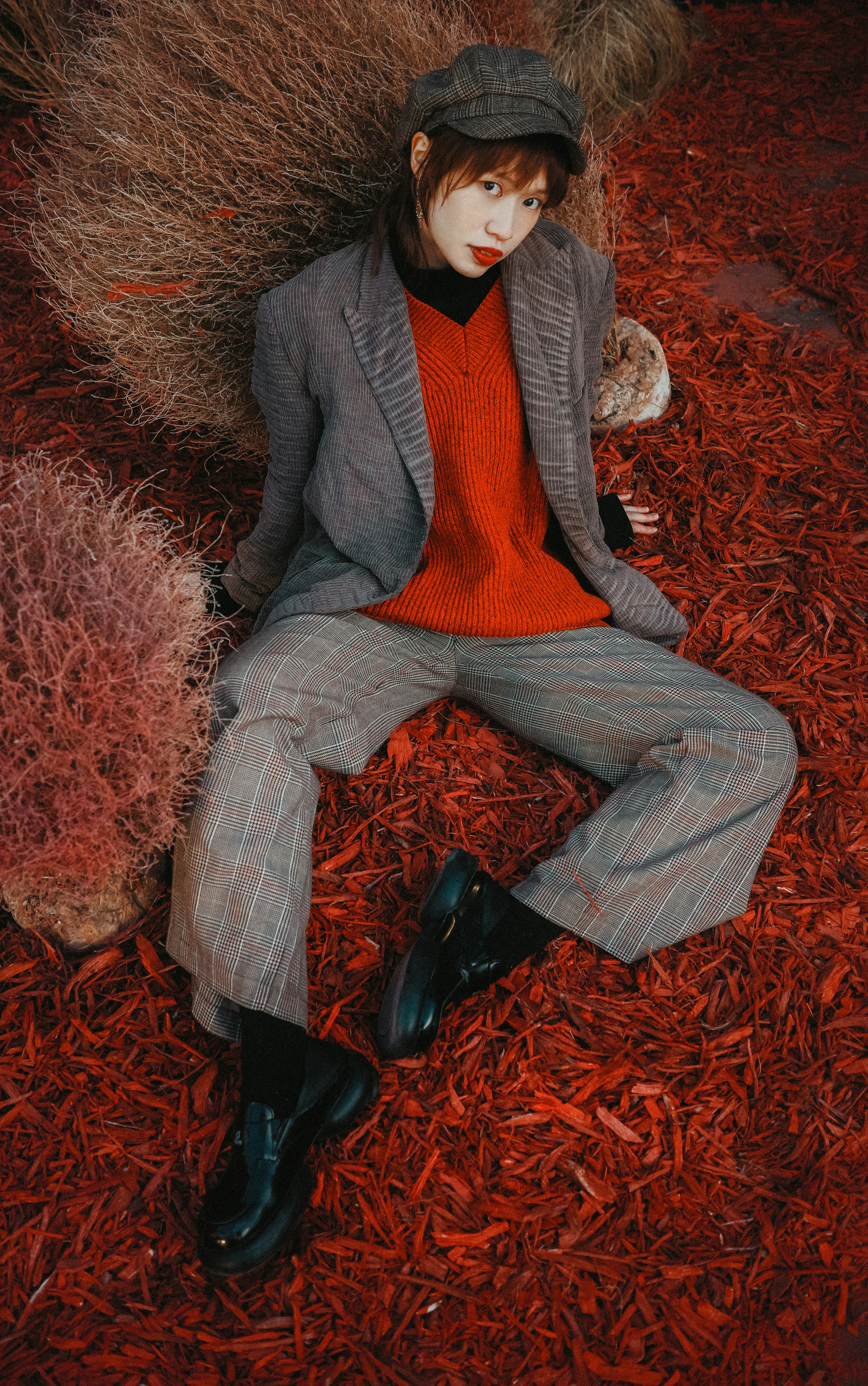 Autumnal-styled portrait with a model seated among red foliage, wearing a gray suit, orange knit sweater, and cap; a fashion-forward, editorial vibe with emphasis on texture and color contrast.