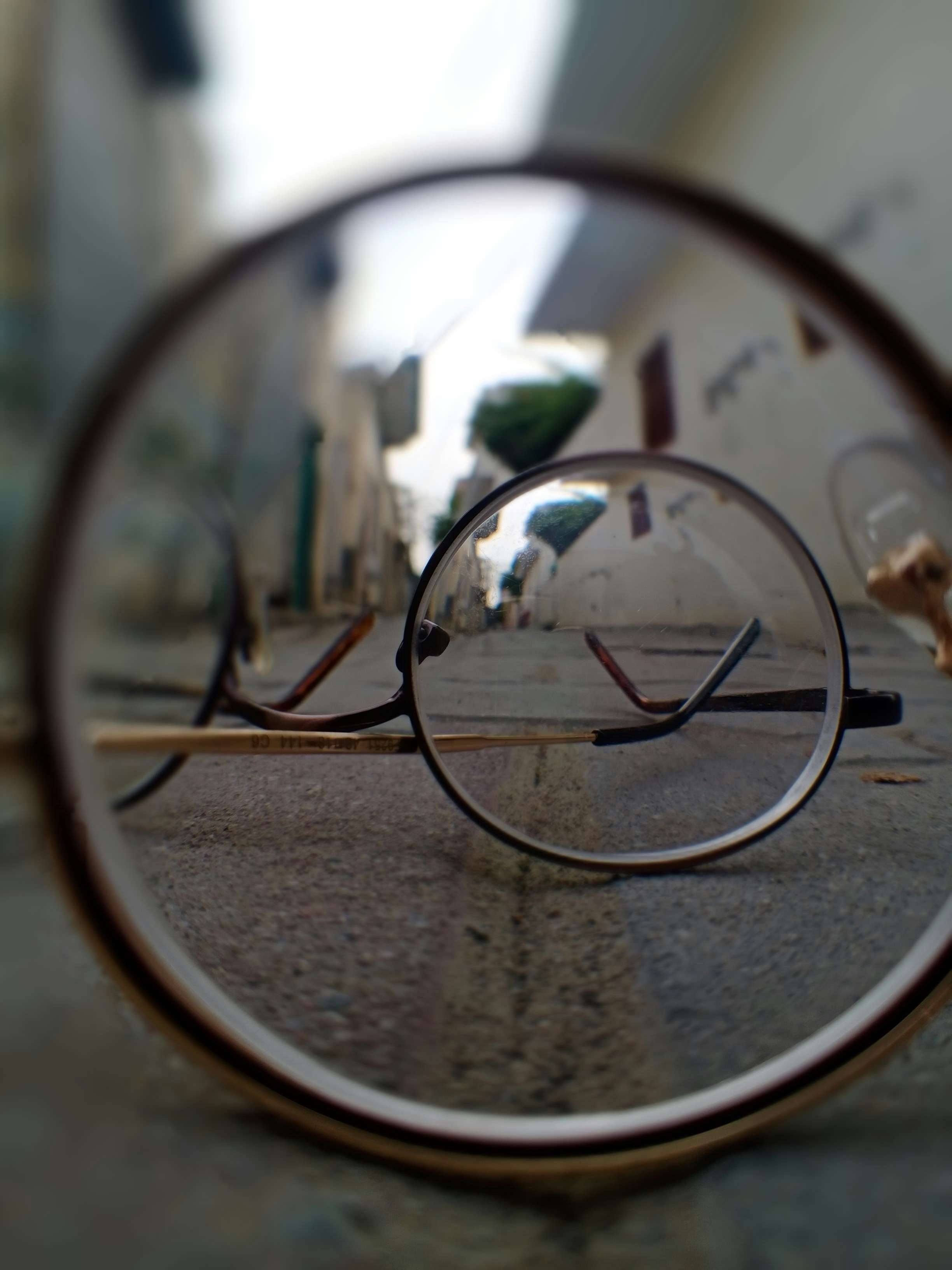 Close-up of a pair of round eyeglasses resting on a textured street, with the foreground ring in sharp focus and the alley receding into soft blur. The image emphasizes depth of field and the reflective surfaces.