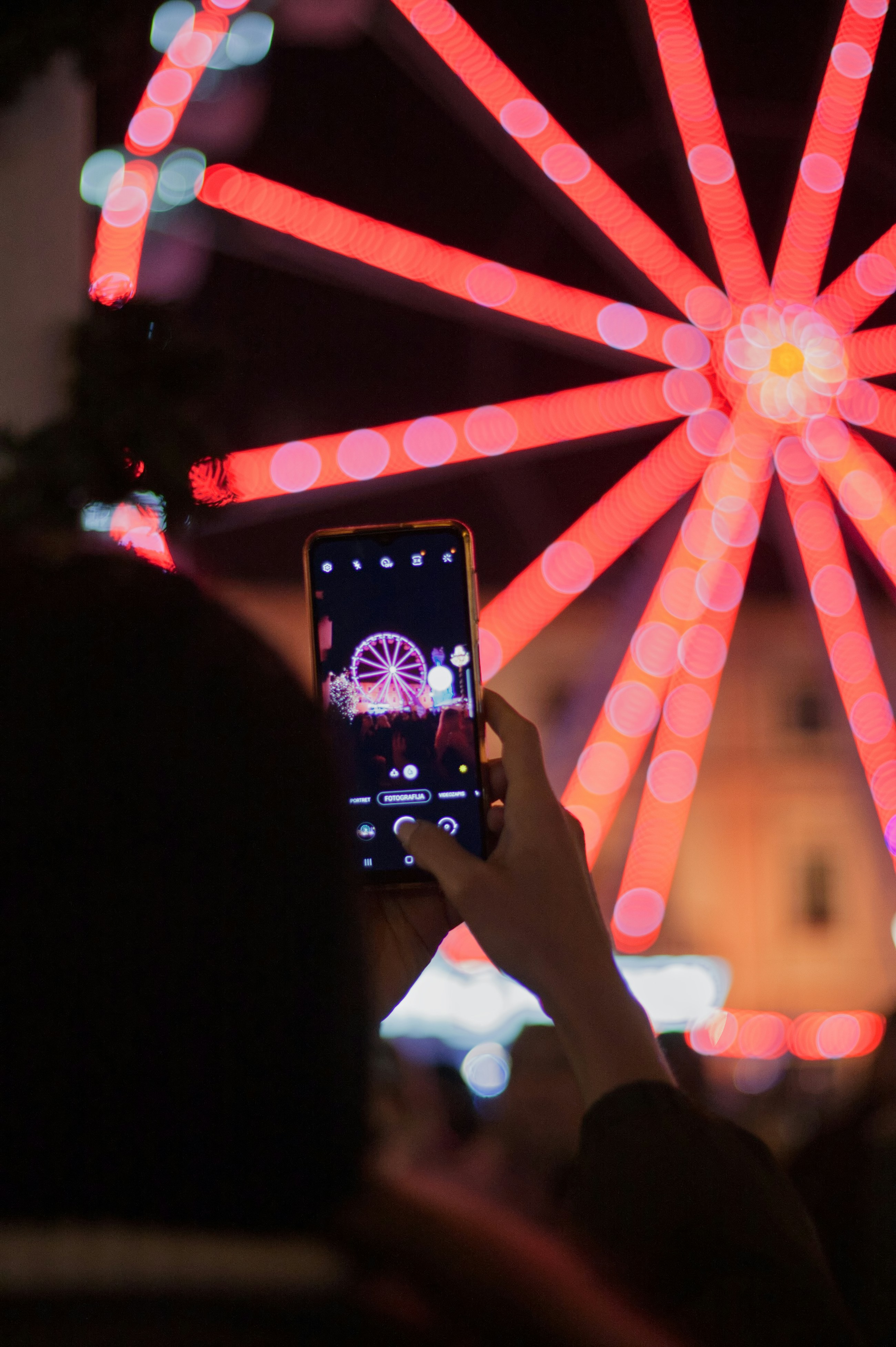 Night scene of a person holding a smartphone to capture a brightly lit Ferris wheel. The device screen shows the wheel amid red bokeh spokes.
