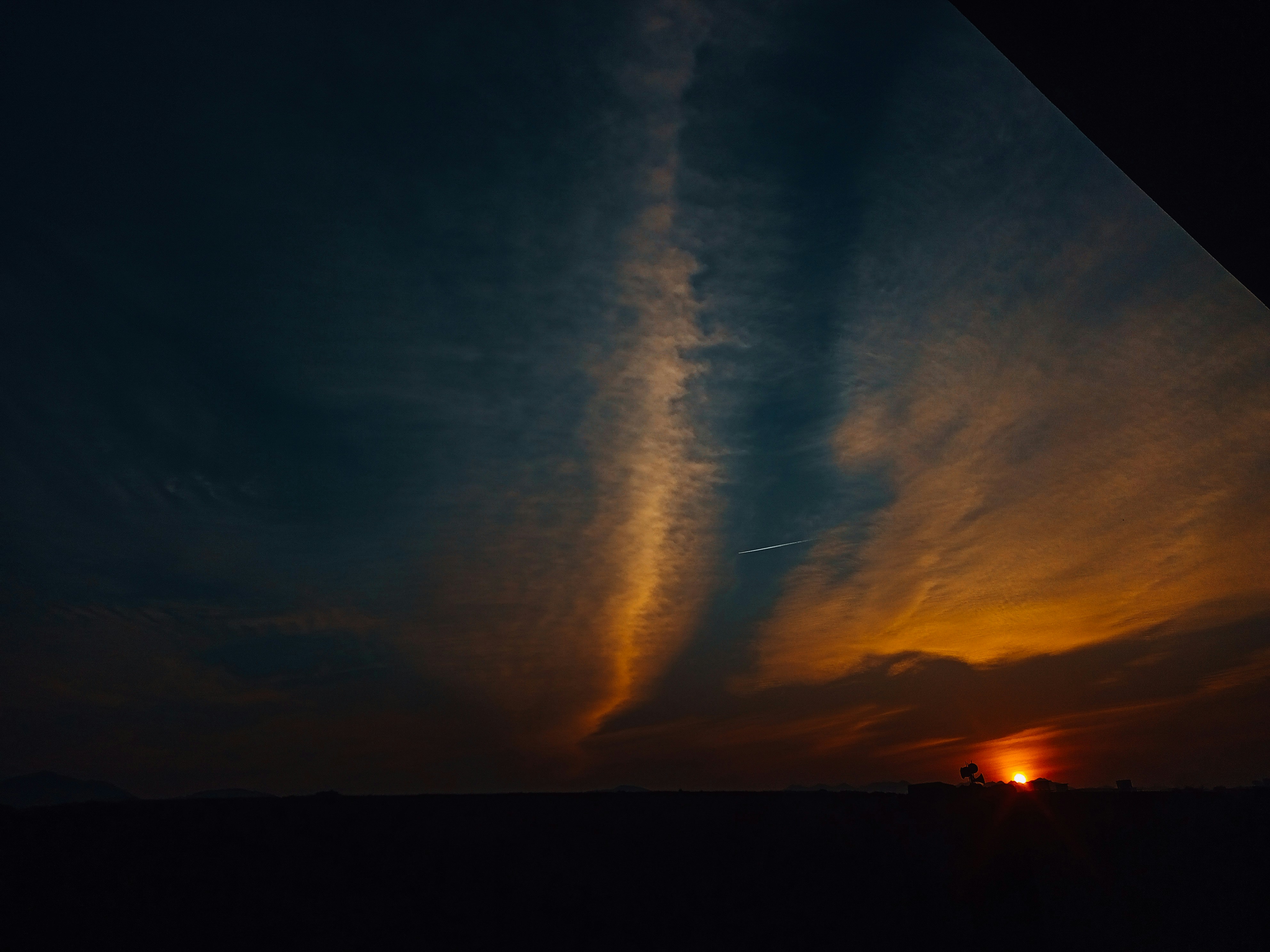 Sunset over a flat horizon with a bright contrail illuminated by the setting sun, painting a warm sky above a dark silhouette.