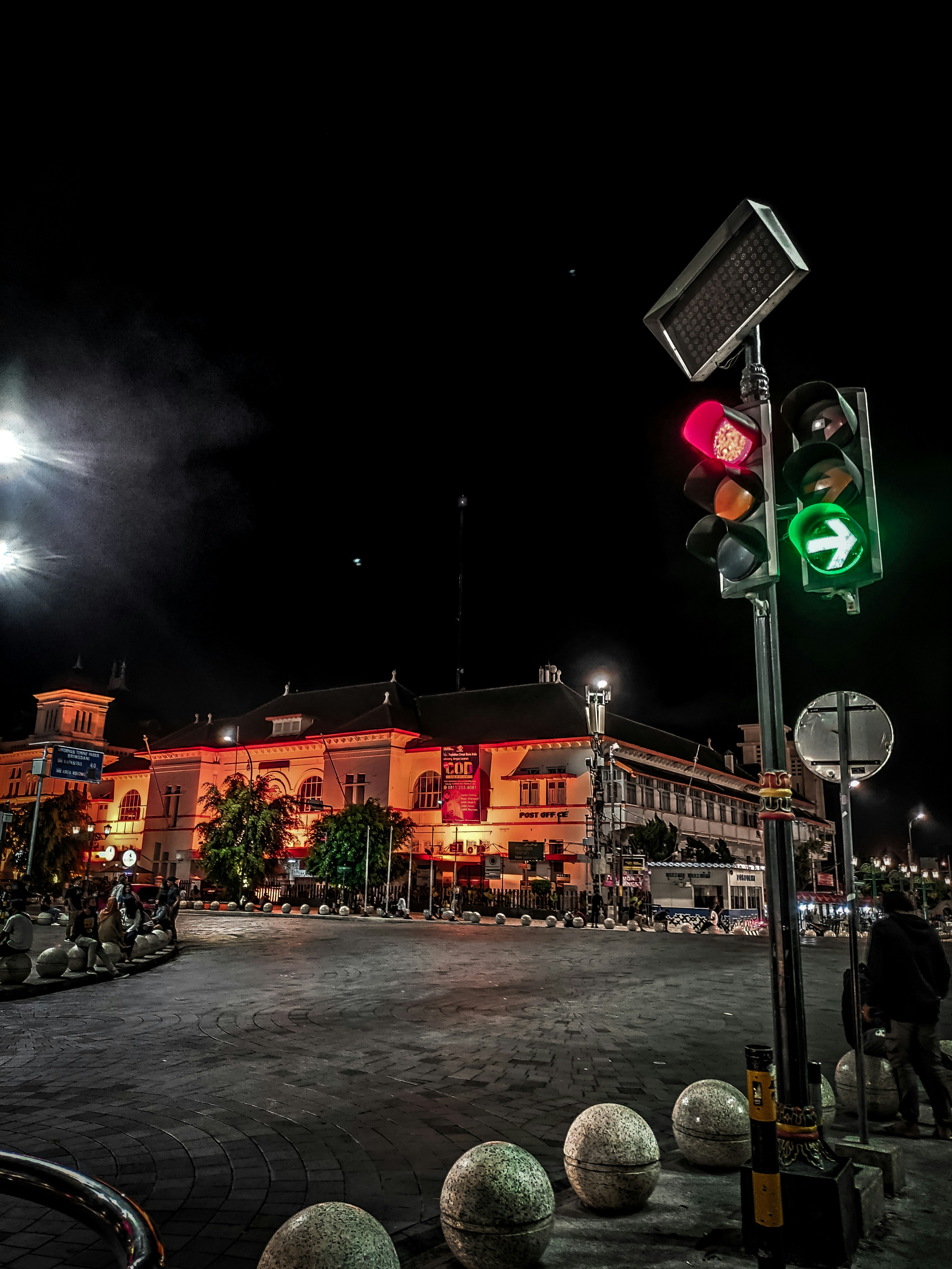 Nighttime city square featuring an amber-lit historic façade as the focal point, a green pedestrian signal nearby, and scattered pedestrians.