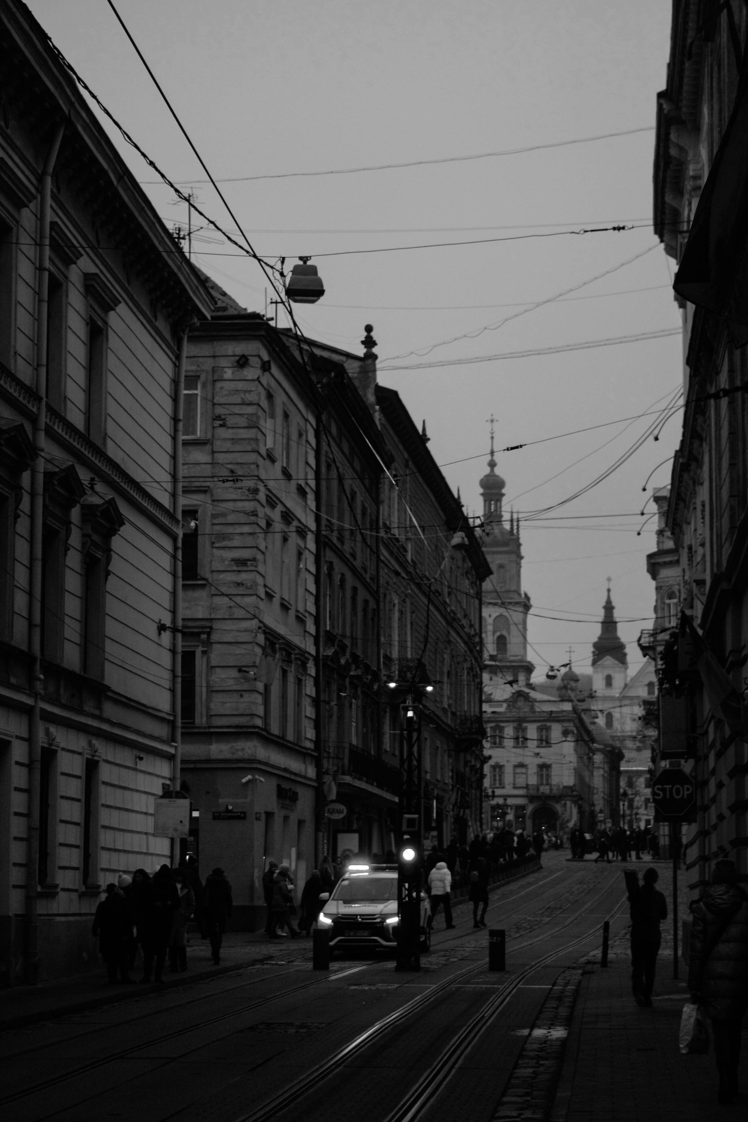 Narrow street lined with historic buildings, showcasing a blend of architecture under a muted sky. A lone car navigates the cobblestone path amidst pedestrians.