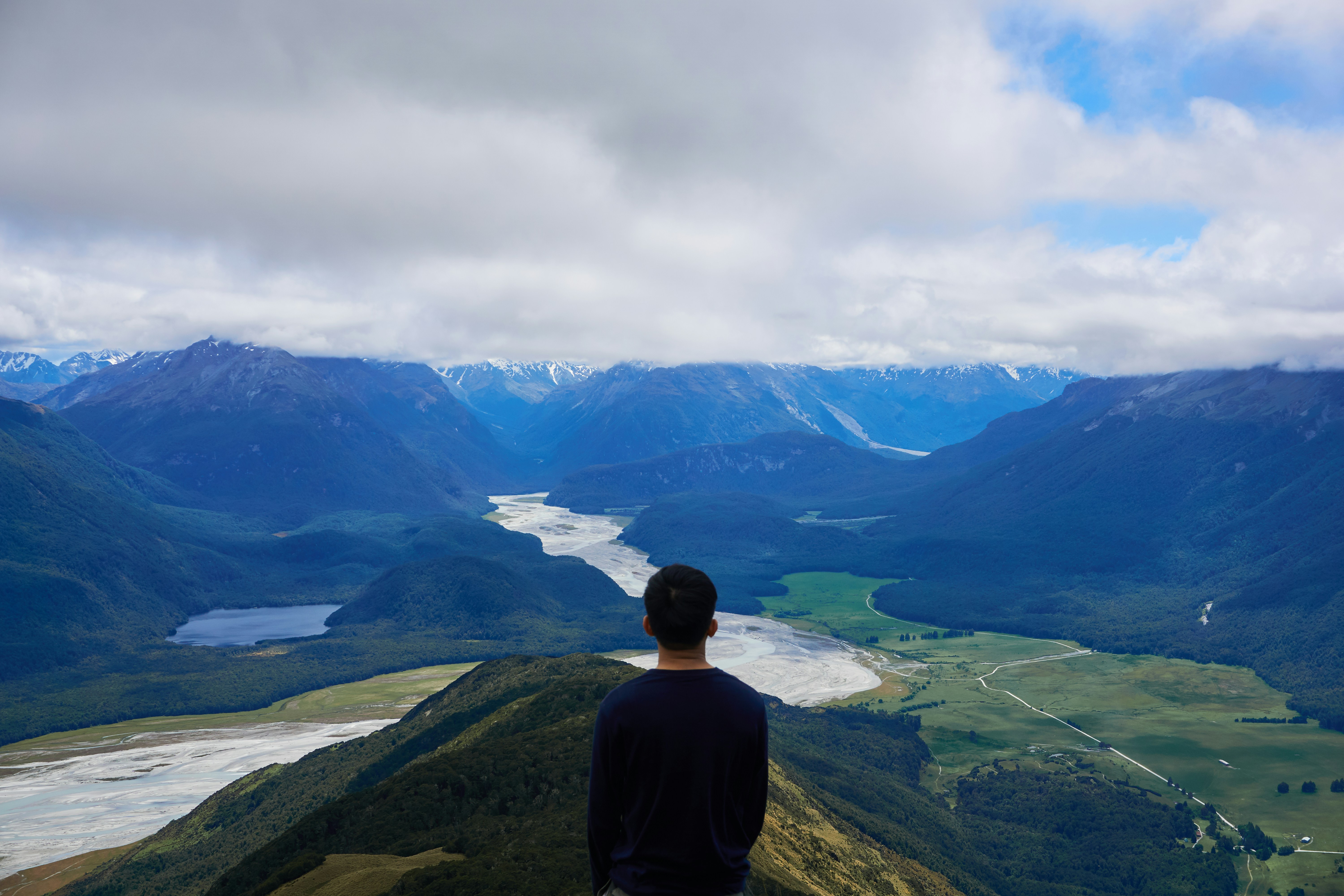 Person standing on a hill overlooking a sprawling valley with winding rivers and distant mountains under a partly cloudy sky.