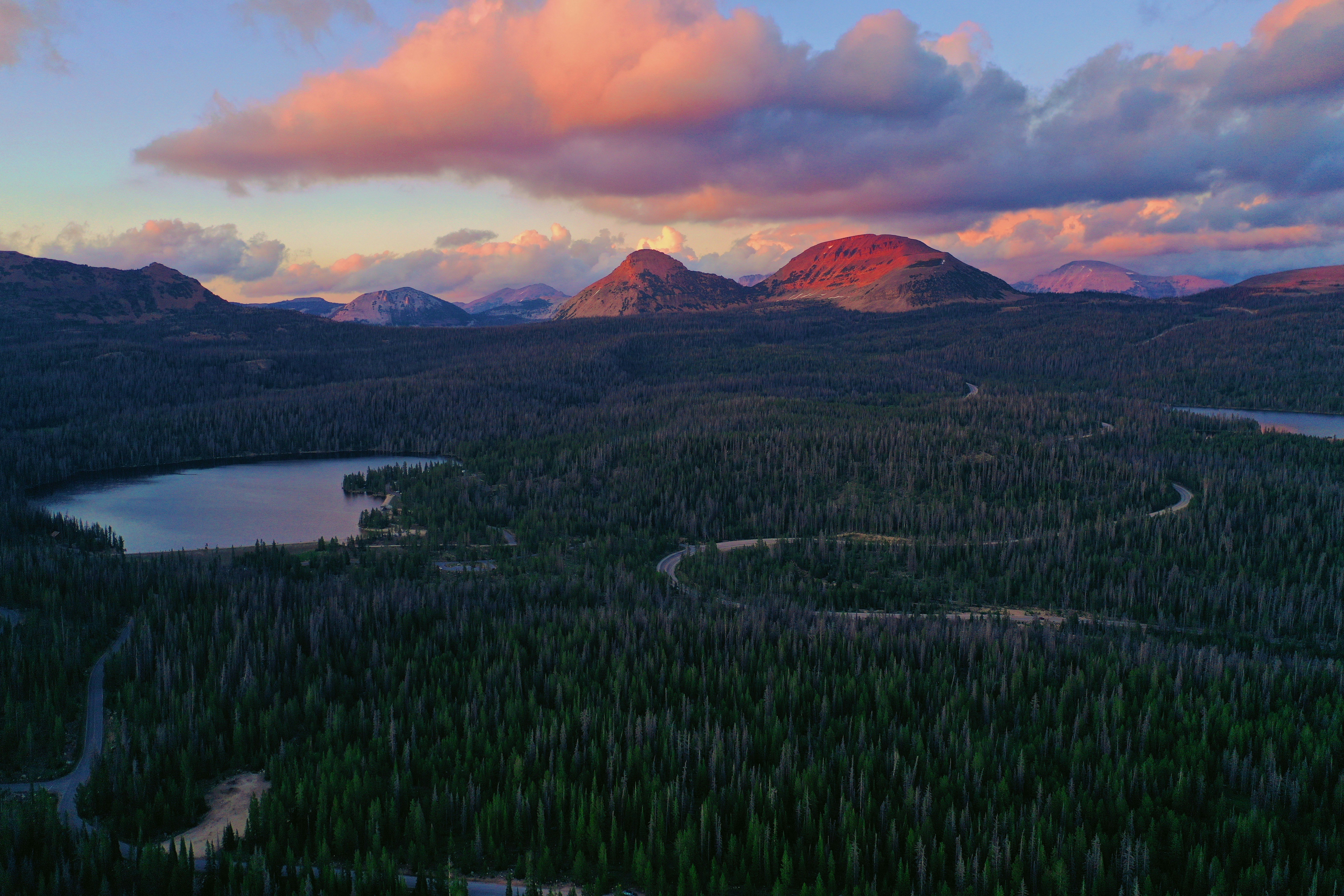 Sunset casts a warm glow over a forested landscape with winding roads and distant mountains.