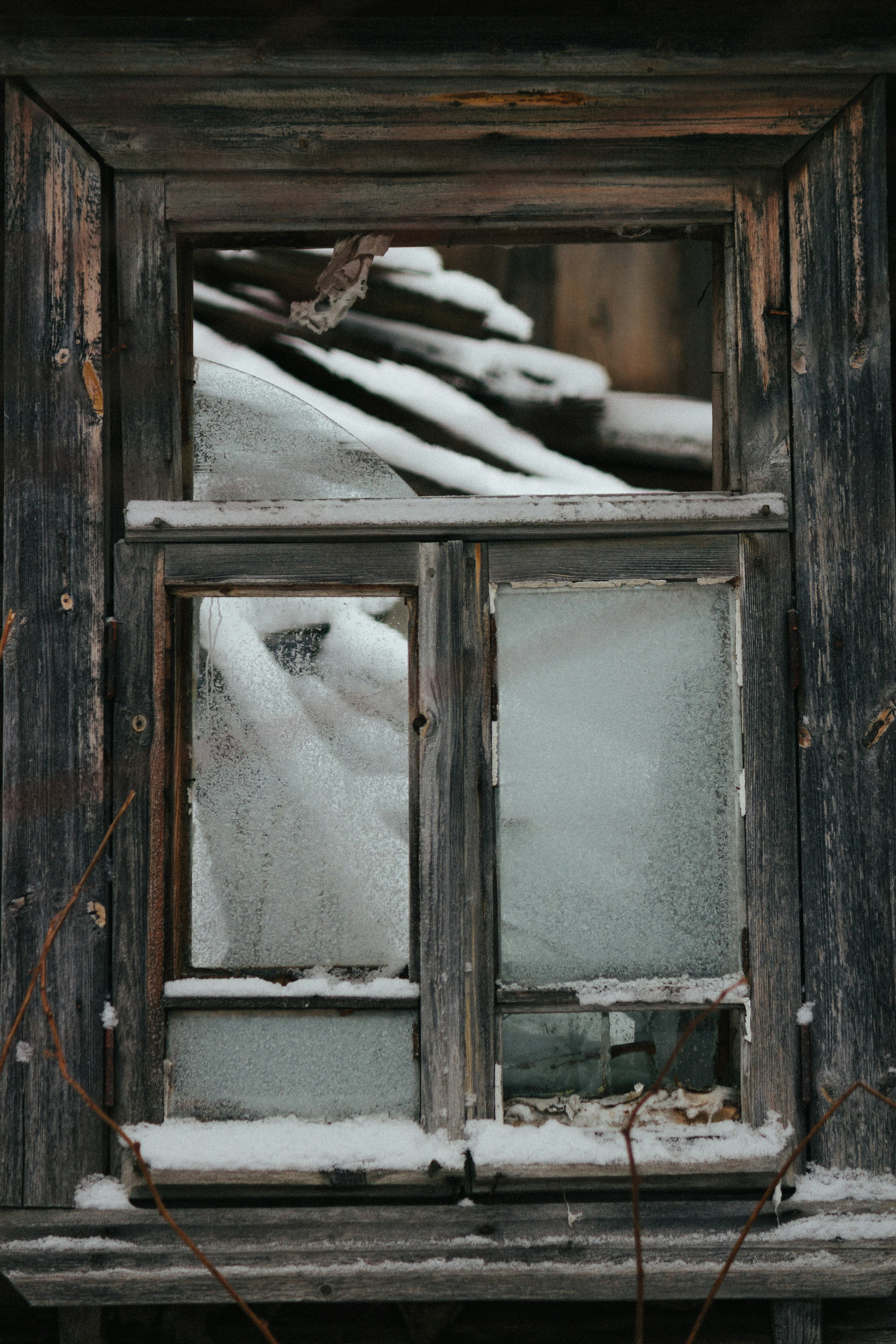 A snow-covered, weathered wooden window frame with frosty glass panes, capturing a glimpse of a snow-laden roof outside.
