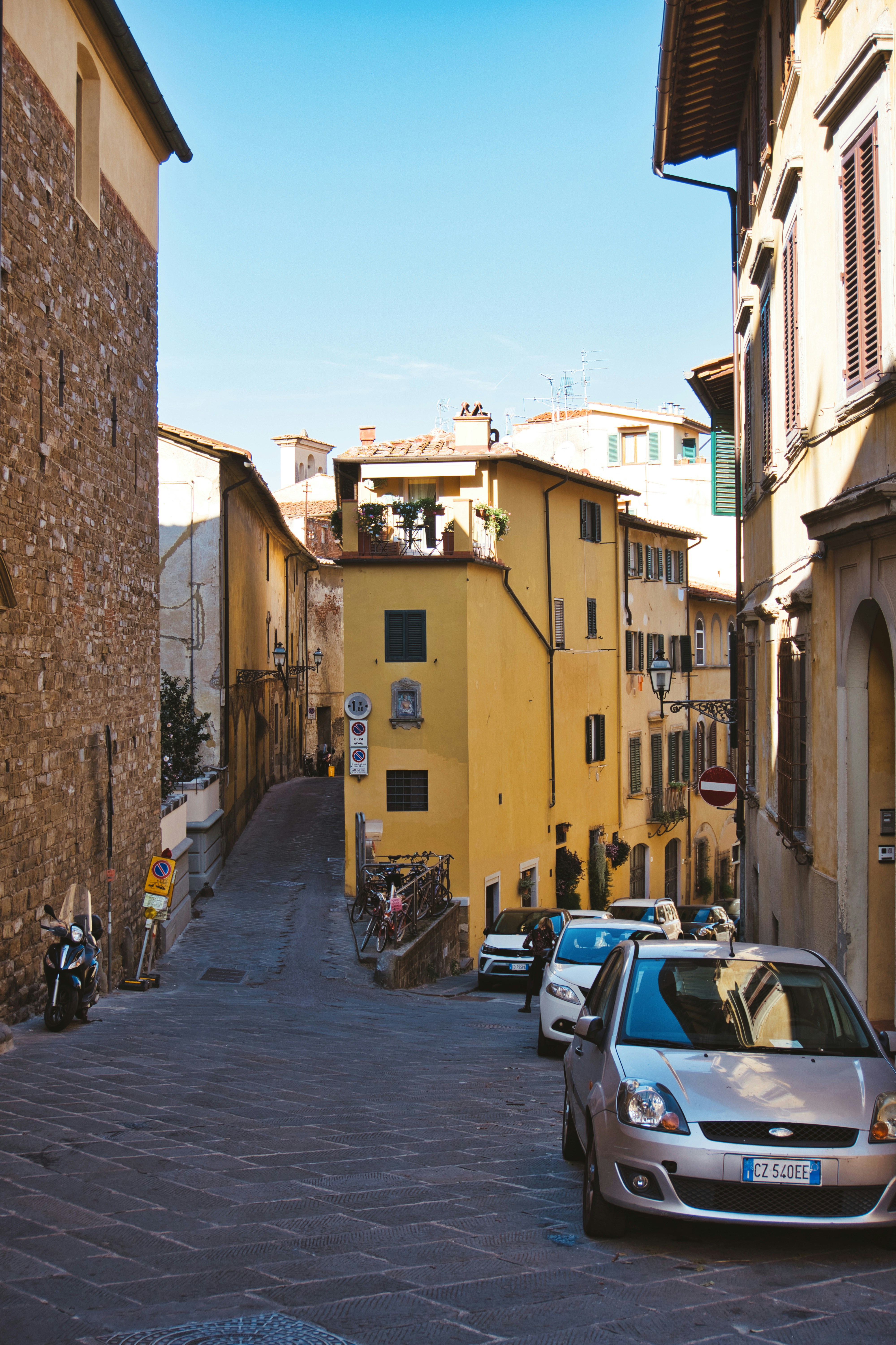 Narrow winding street in a Tuscan town, flanked by rustic buildings and parked cars. The vibrant yellow facade adds a warm touch to the scene.