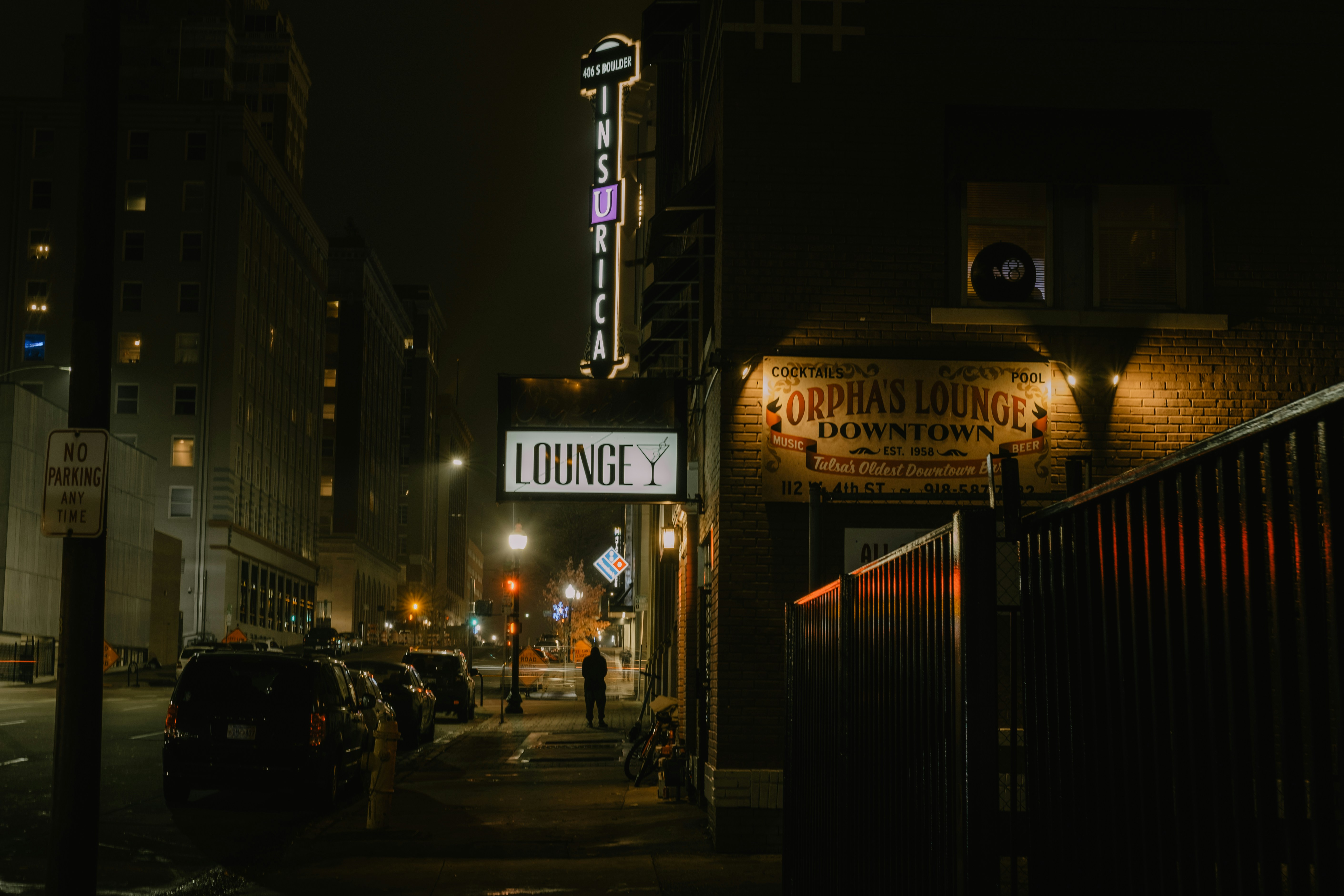 Dimly lit street at night with glowing lounge signs and parked cars lining the road.