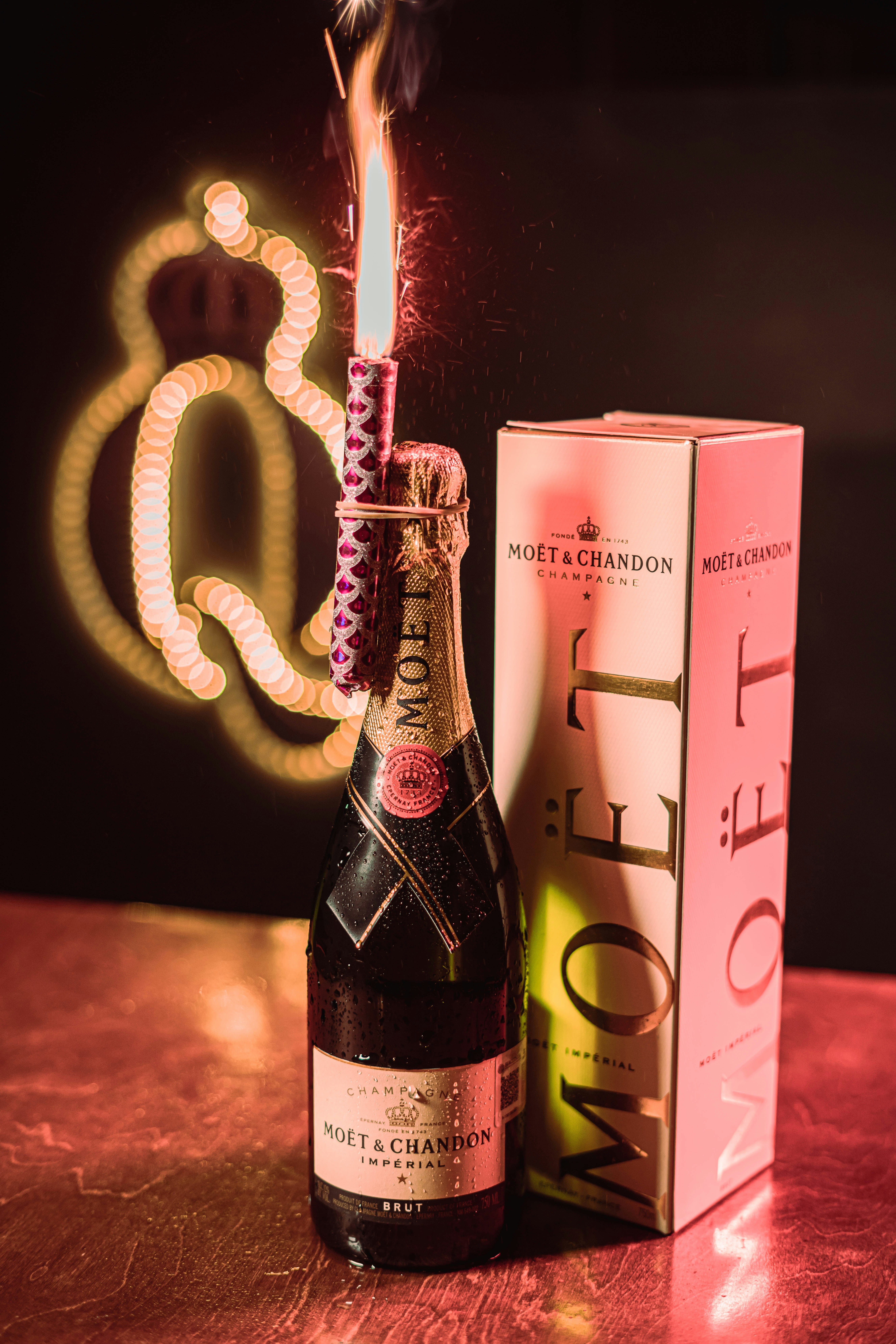 Champagne bottle with fireworks on a reflective surface, illuminated by a glowing crown-shaped neon light.