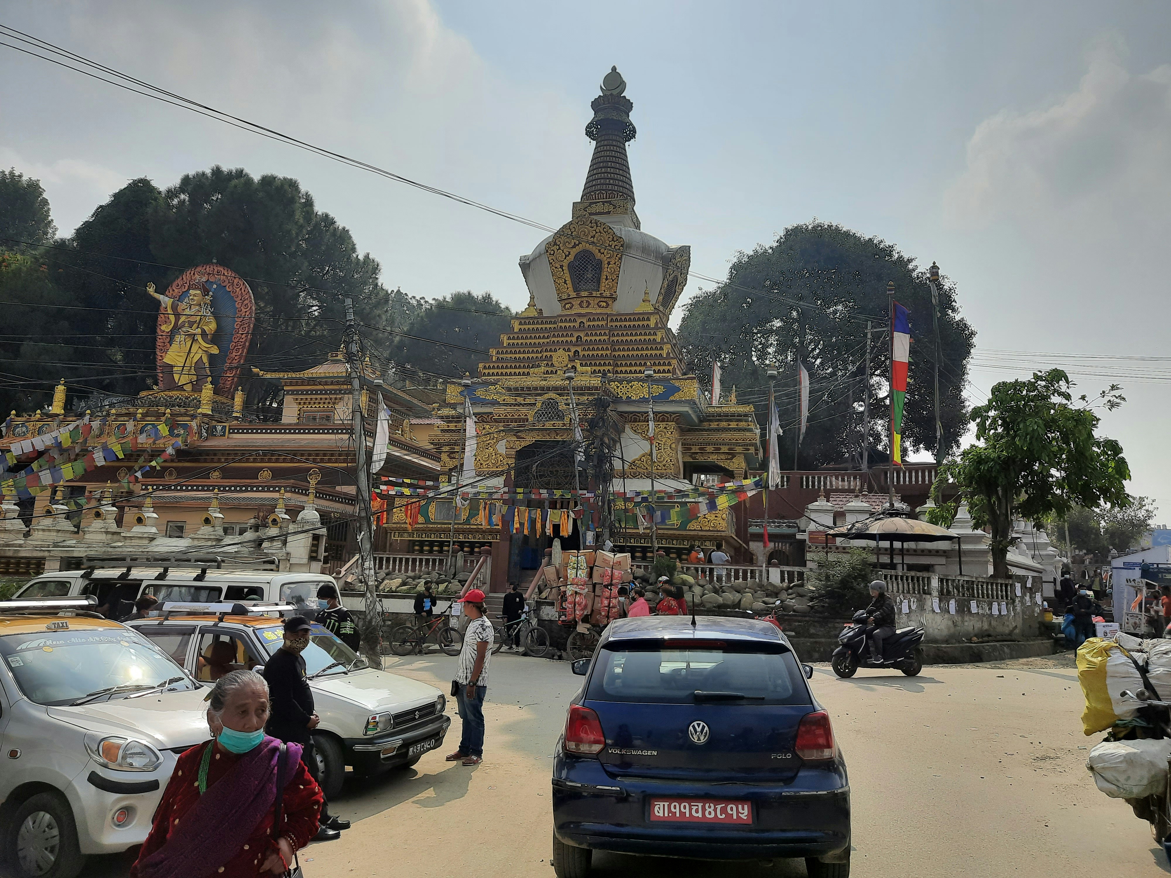 Gilded stupa rises behind a busy street, framed by colorful prayer flags and parked cars around the temple precinct.