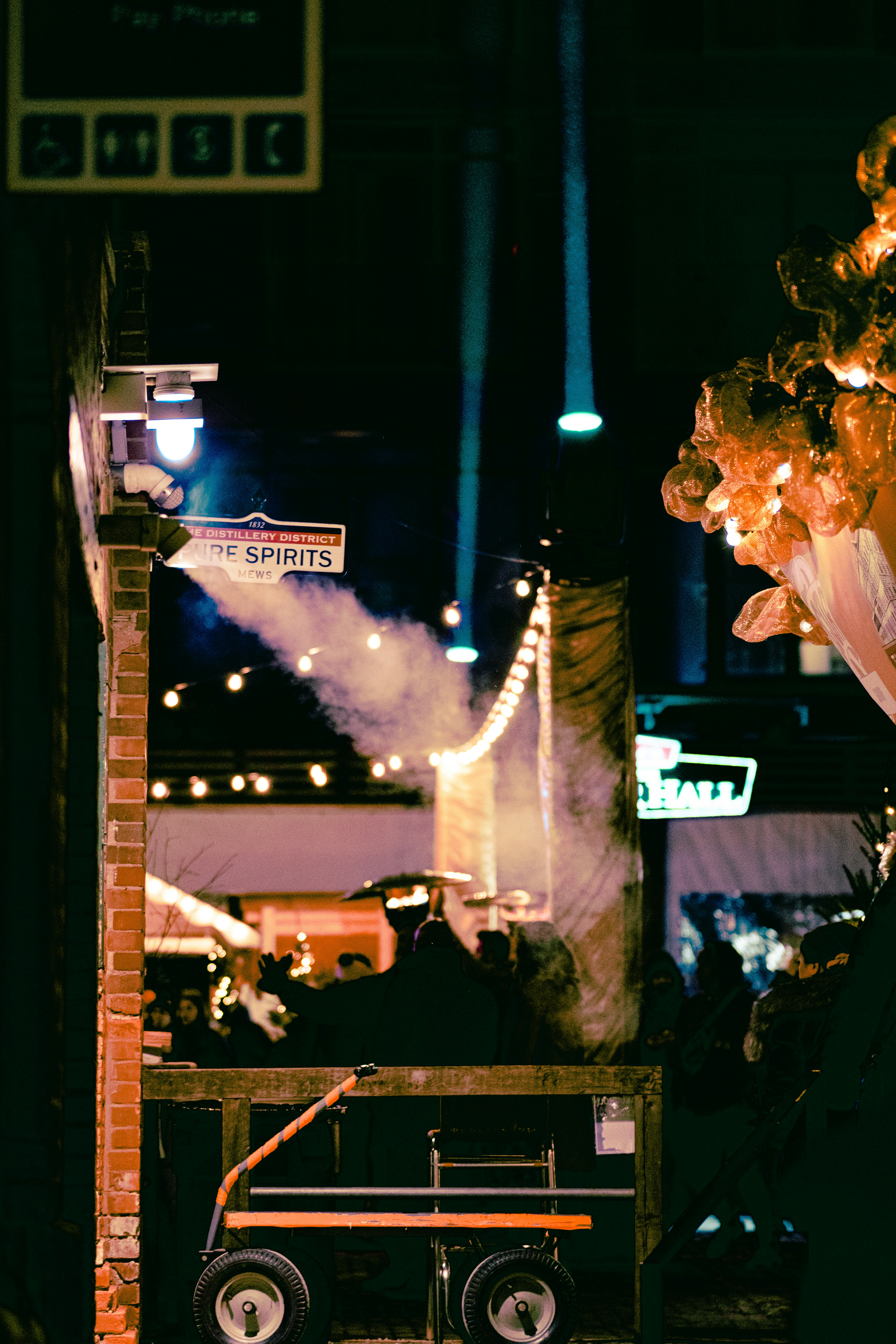 Warm lights illuminate a bustling scene in a distillery district, with steam rising from a cart and people mingling in the background.