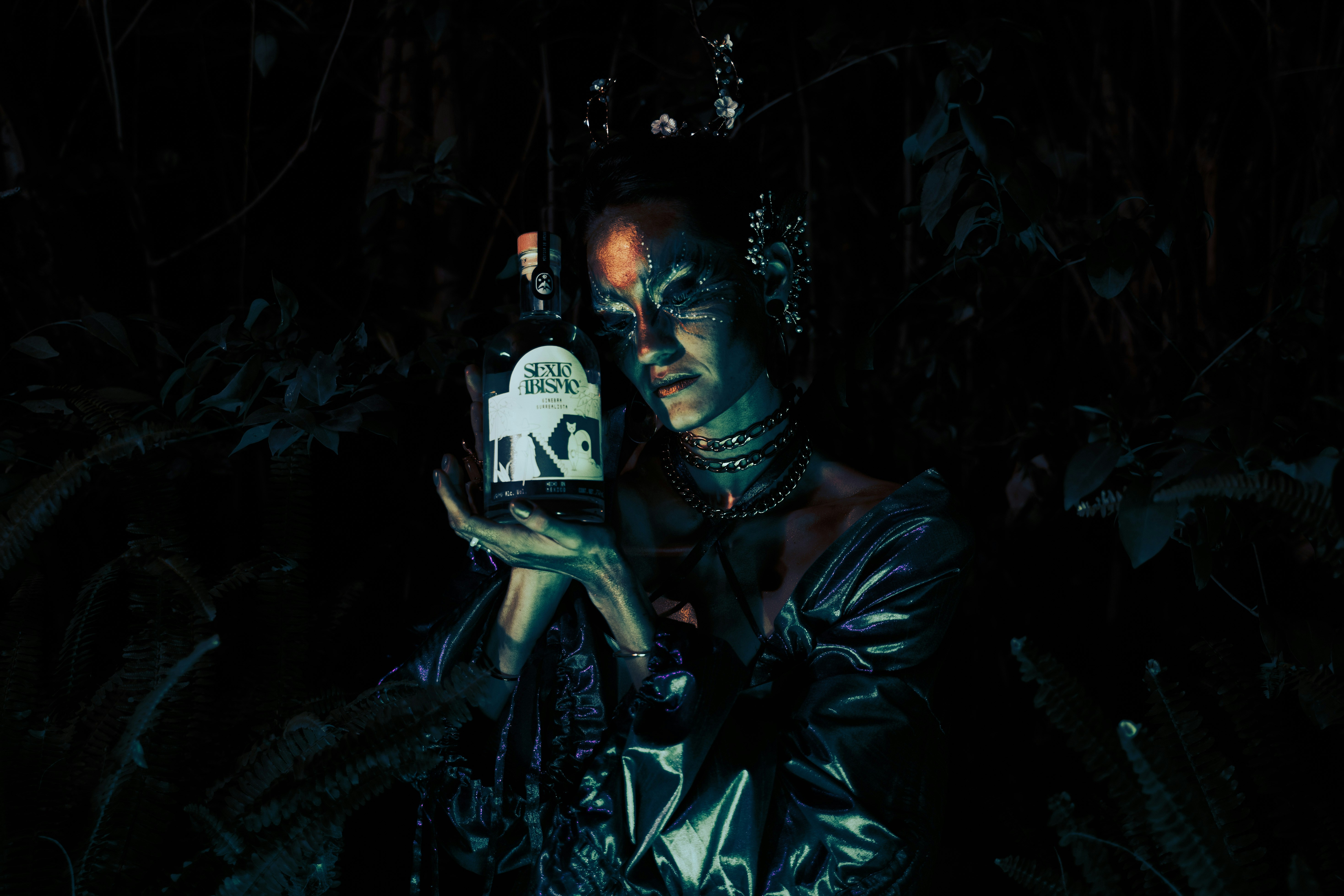 Person holding a bottle of gin amidst dark foliage, illuminated by subtle lighting.