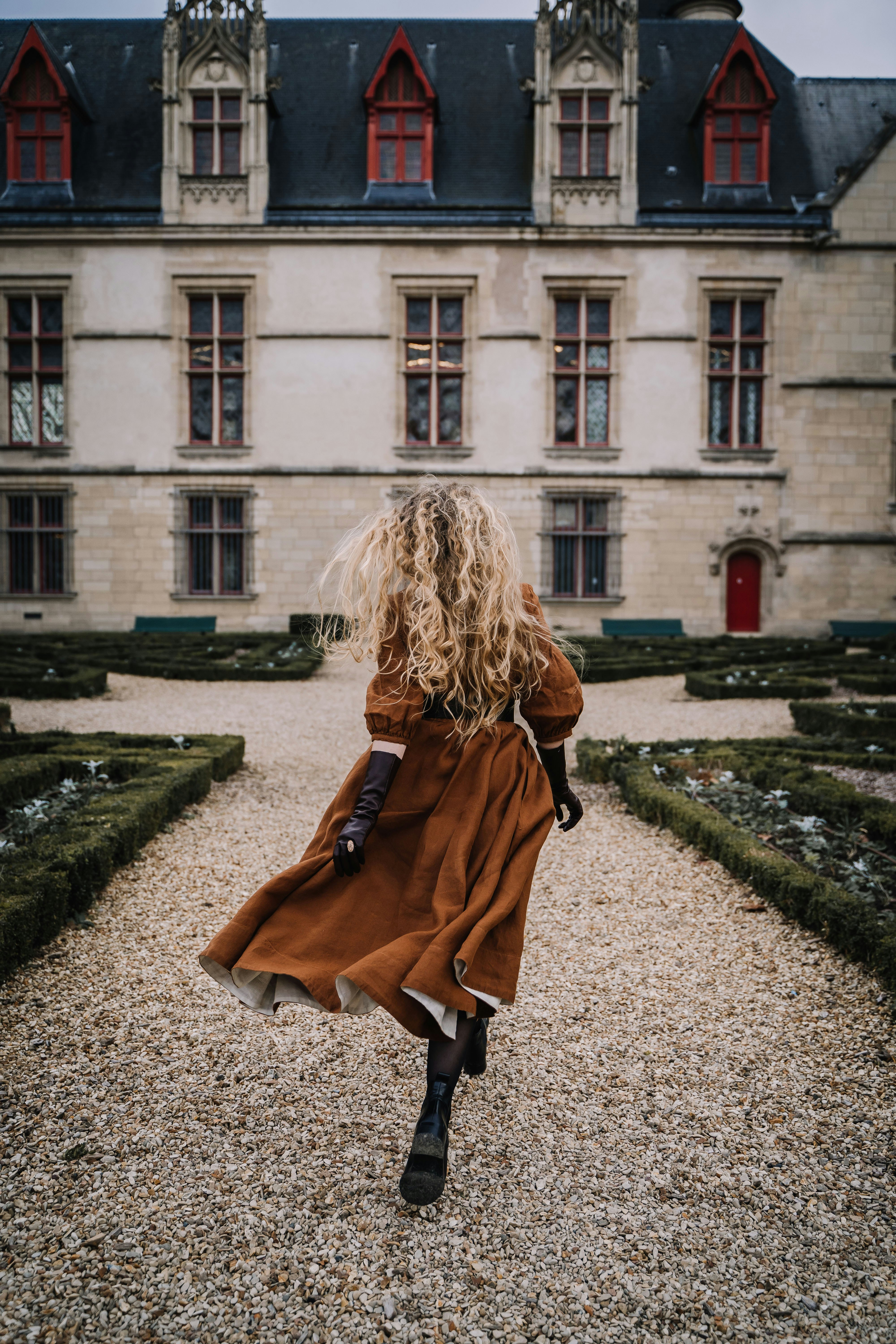 A girl in a rust-colored dress runs down a gravel path between formal hedges toward an old stone building with red-trimmed windows, captured in soft daylight. The scene resembles a staged setting, with the figure as the focal point amid the historic architecture.